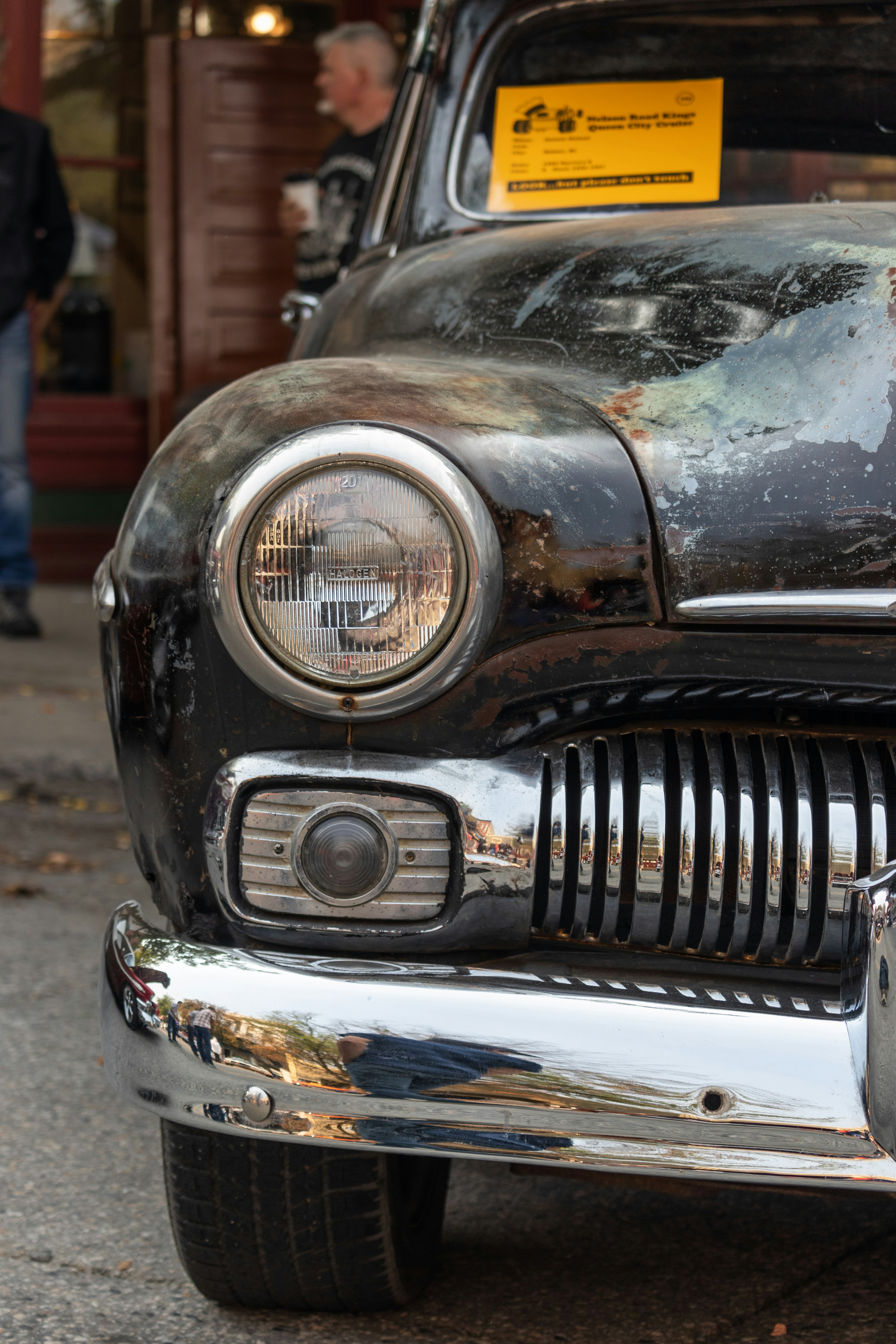 Close-up view of a vintage car's front end, showcasing its weathered paint and chrome details. A yellow sticker adds a pop of color against the aged surface.