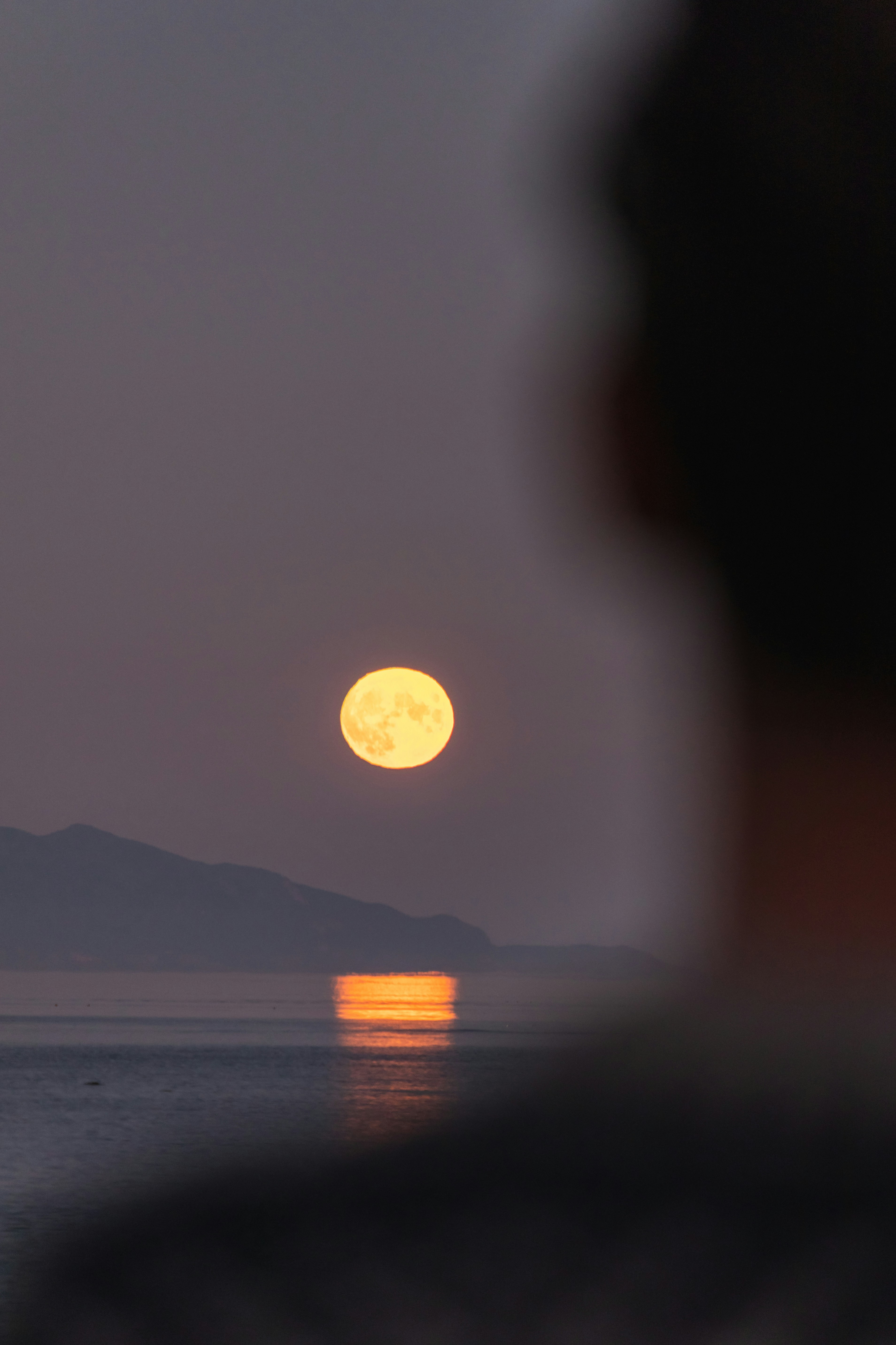 Full moon rising over tranquil waters, casting a golden reflection against distant mountains.