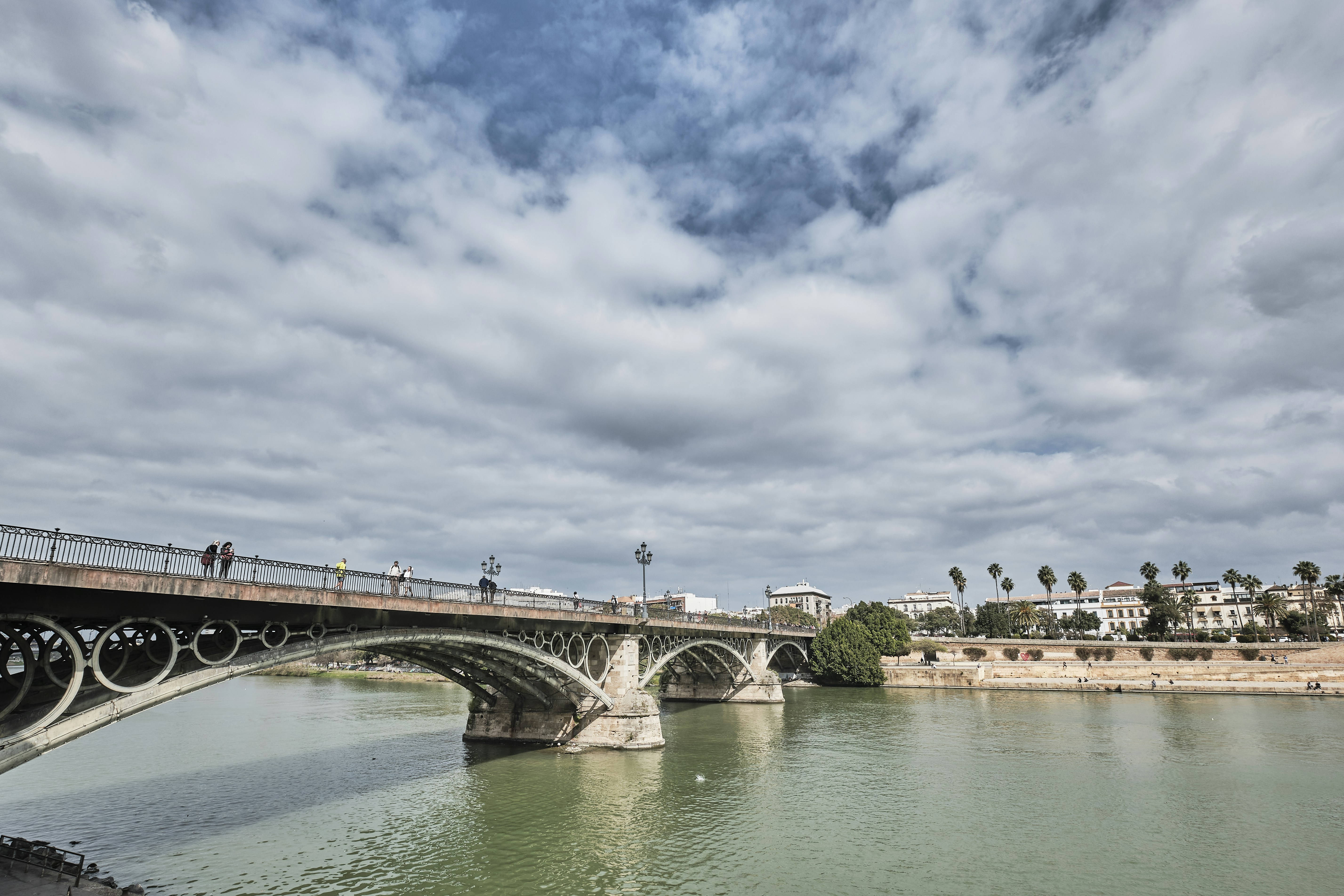 Historic bridge spanning a calm river under a partly cloudy sky, with distant palm trees lining the riverbank.