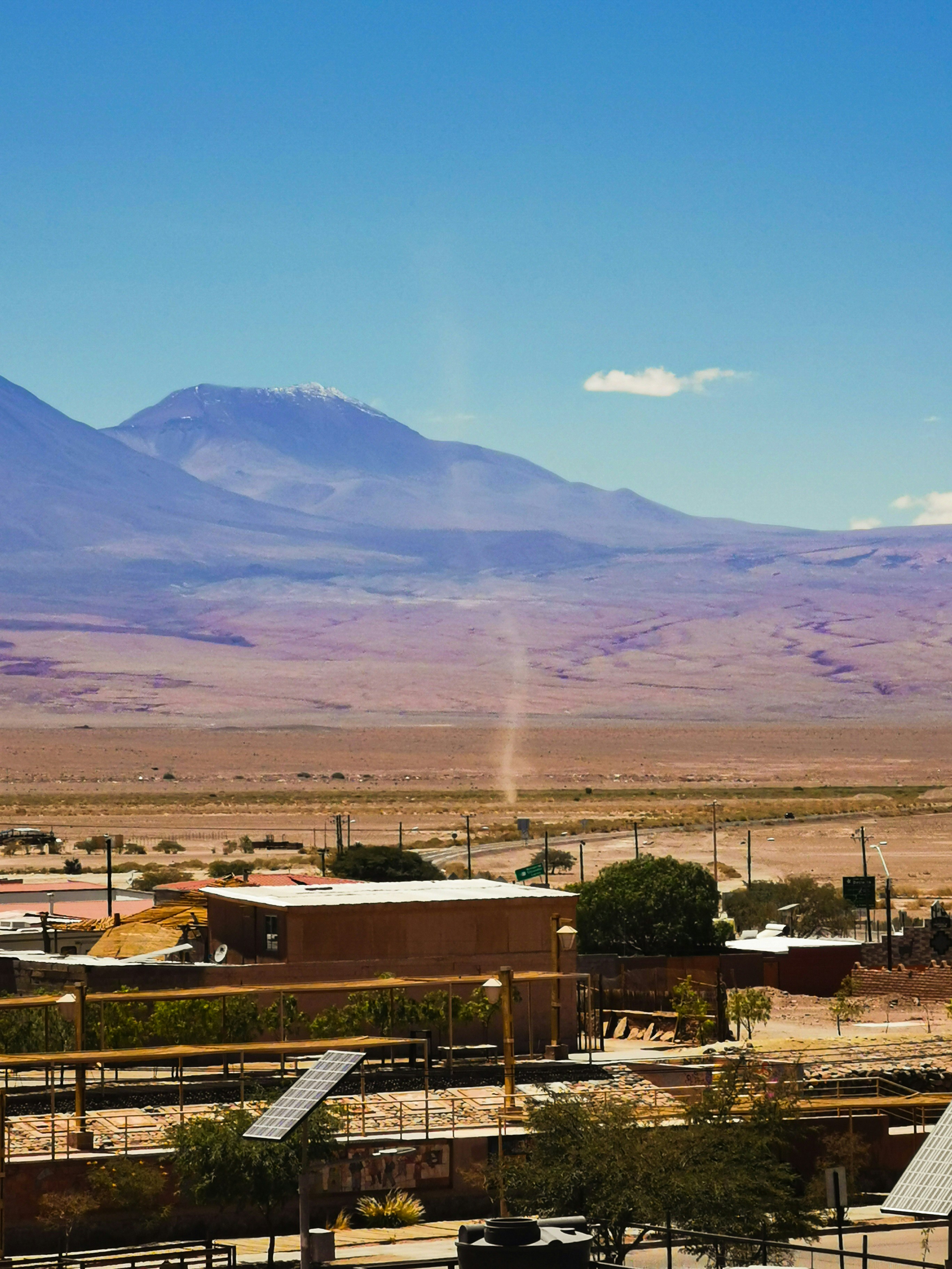 Dust devil spiraling across the arid landscape under a clear blue sky, framed by distant mountains and sparse vegetation.