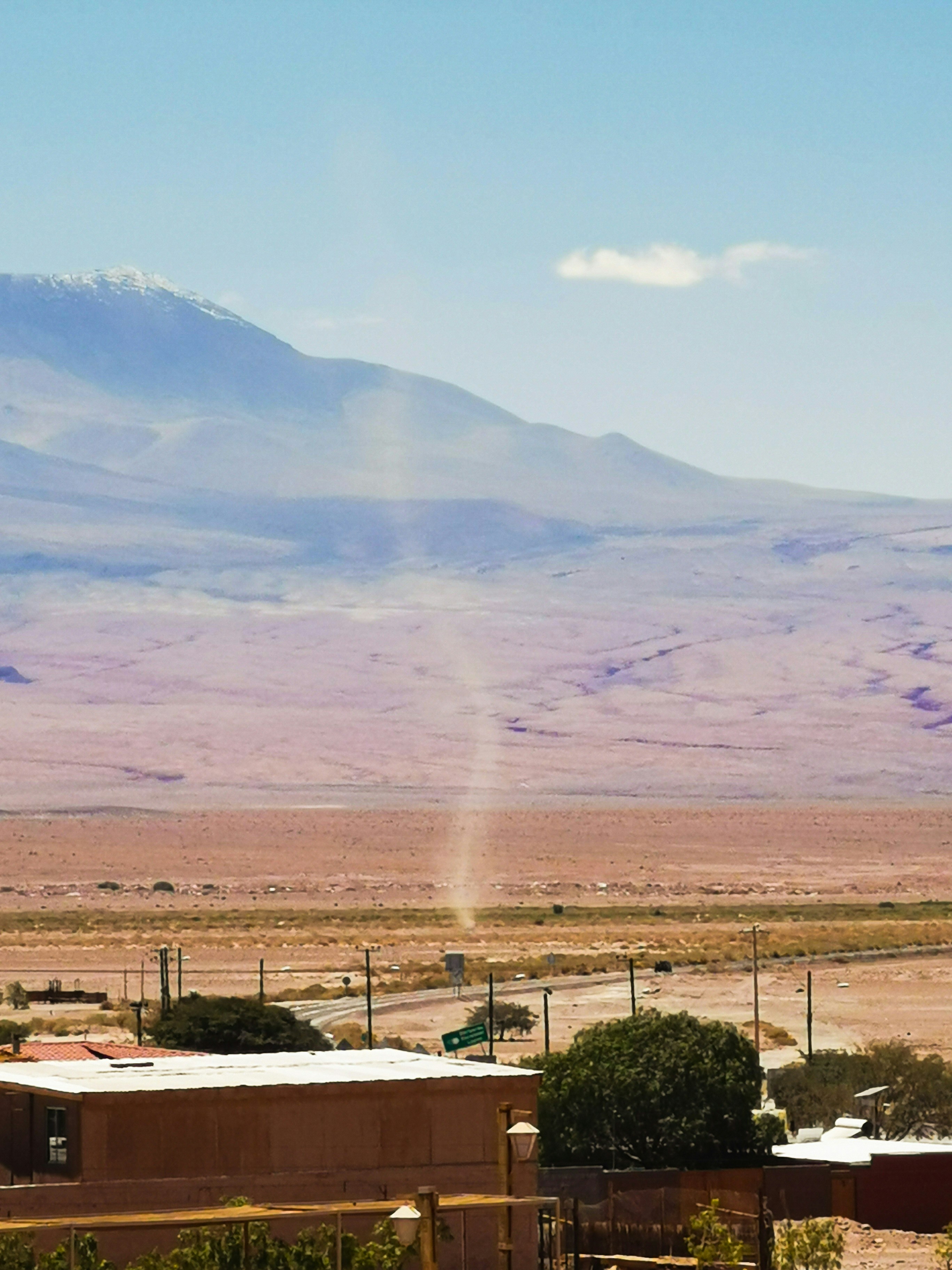 A swirling dust devil rises from the arid landscape, framed by distant mountains under a clear blue sky. The scene captures the essence of a dry, vibrant desert environment.