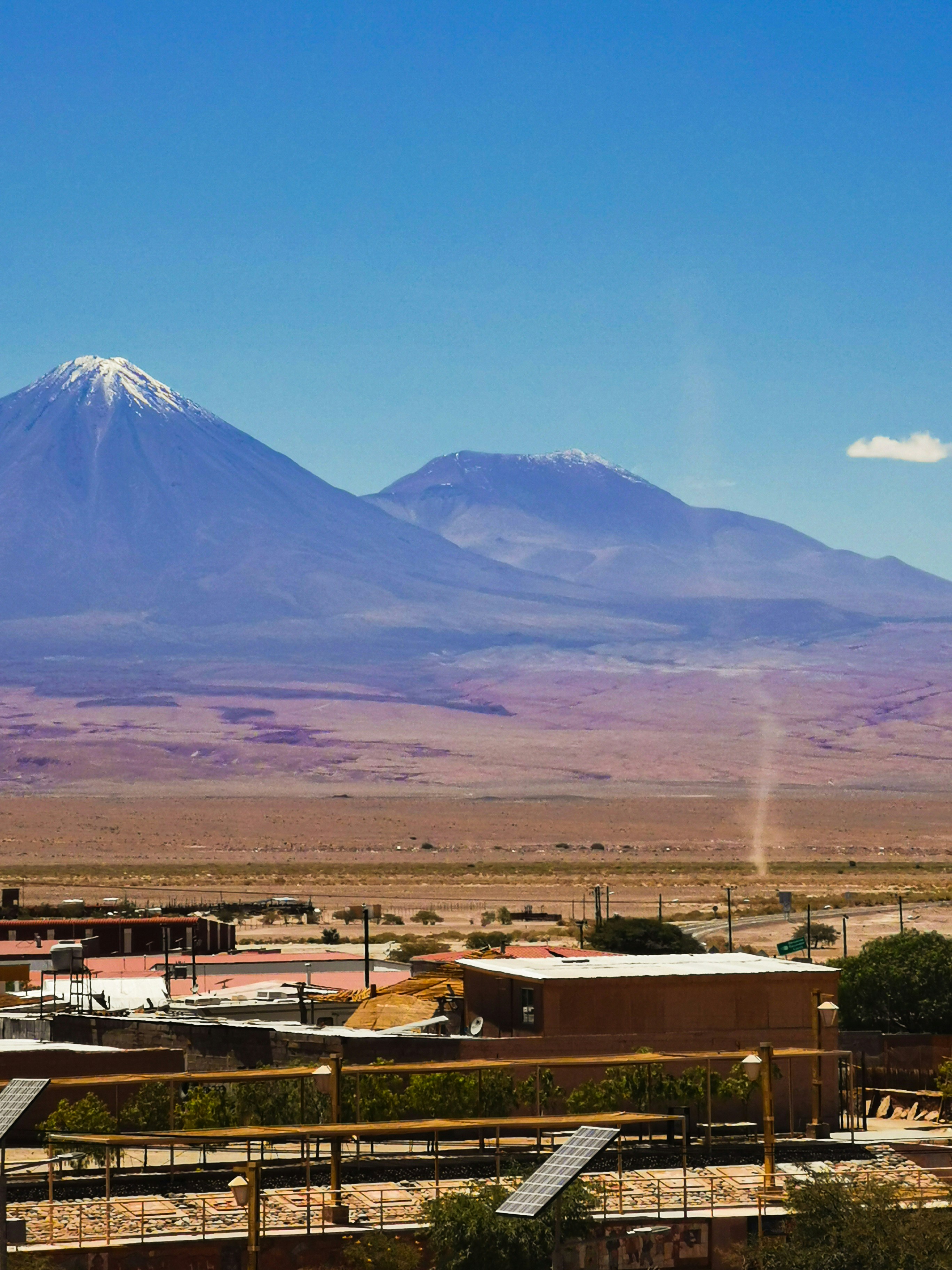Desert town sprawls along the foothills with low buildings and solar panels under a bright blue sky. Snow-capped mountains rise in the background, creating a stark, expansive landscape.