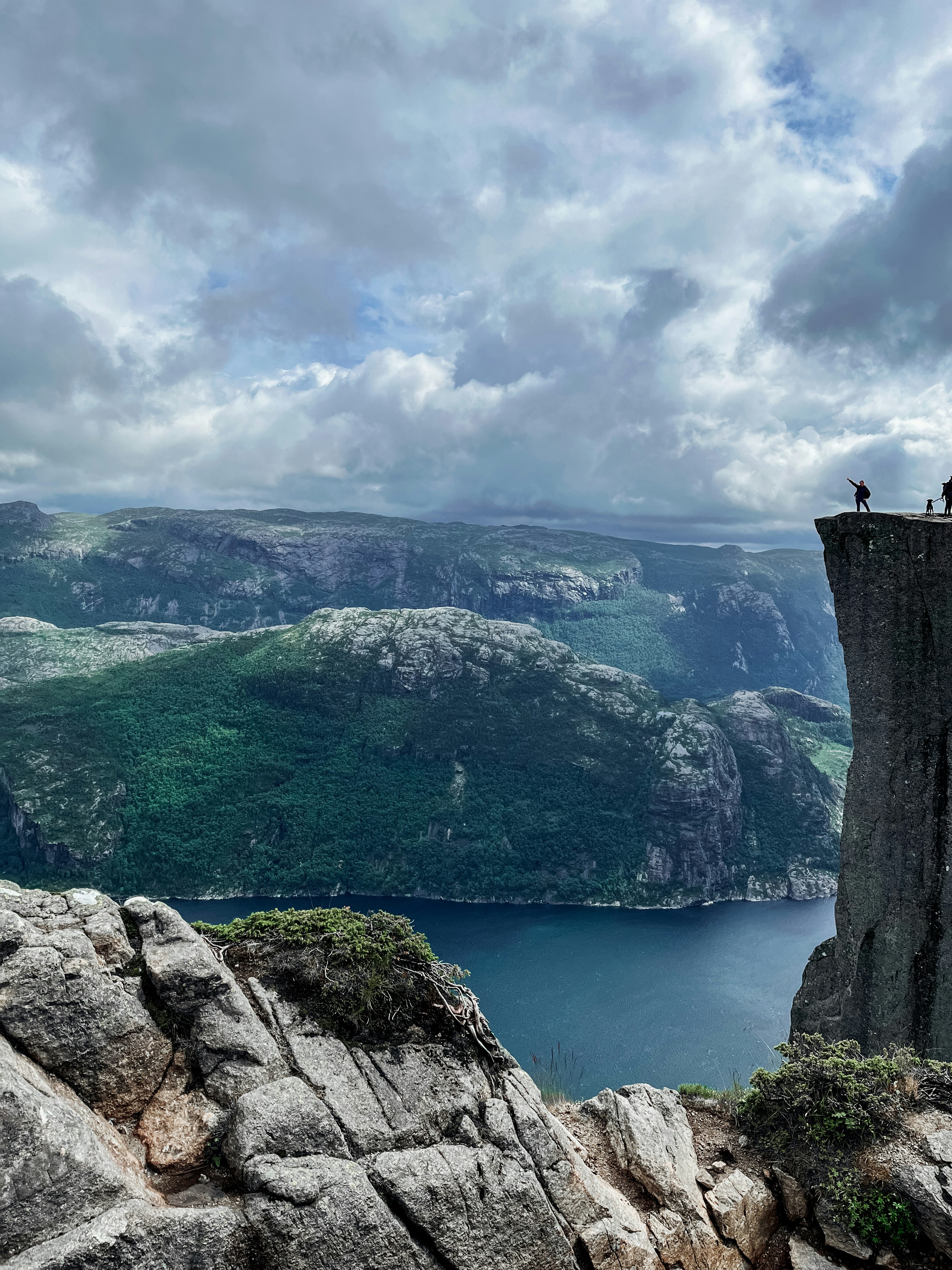 A lone figure stands on a dramatic cliff edge overlooking a vast fjord surrounded by lush greenery and rugged mountains. The sky is filled with dynamic clouds, enhancing the sense of adventure.