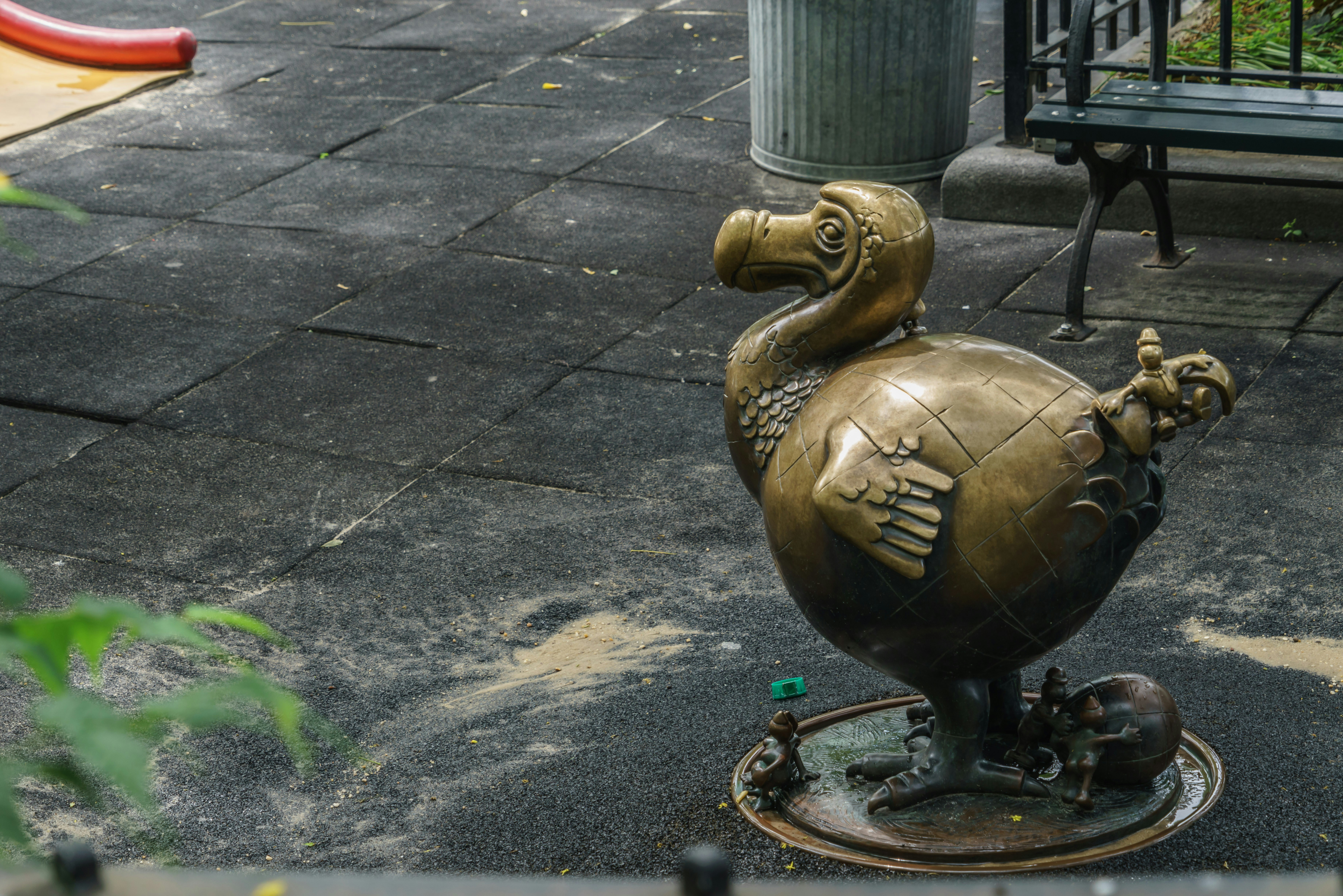Bronze sculpture of a dodo bird perched on a circular base in a playground setting, surrounded by textured pavement and greenery.