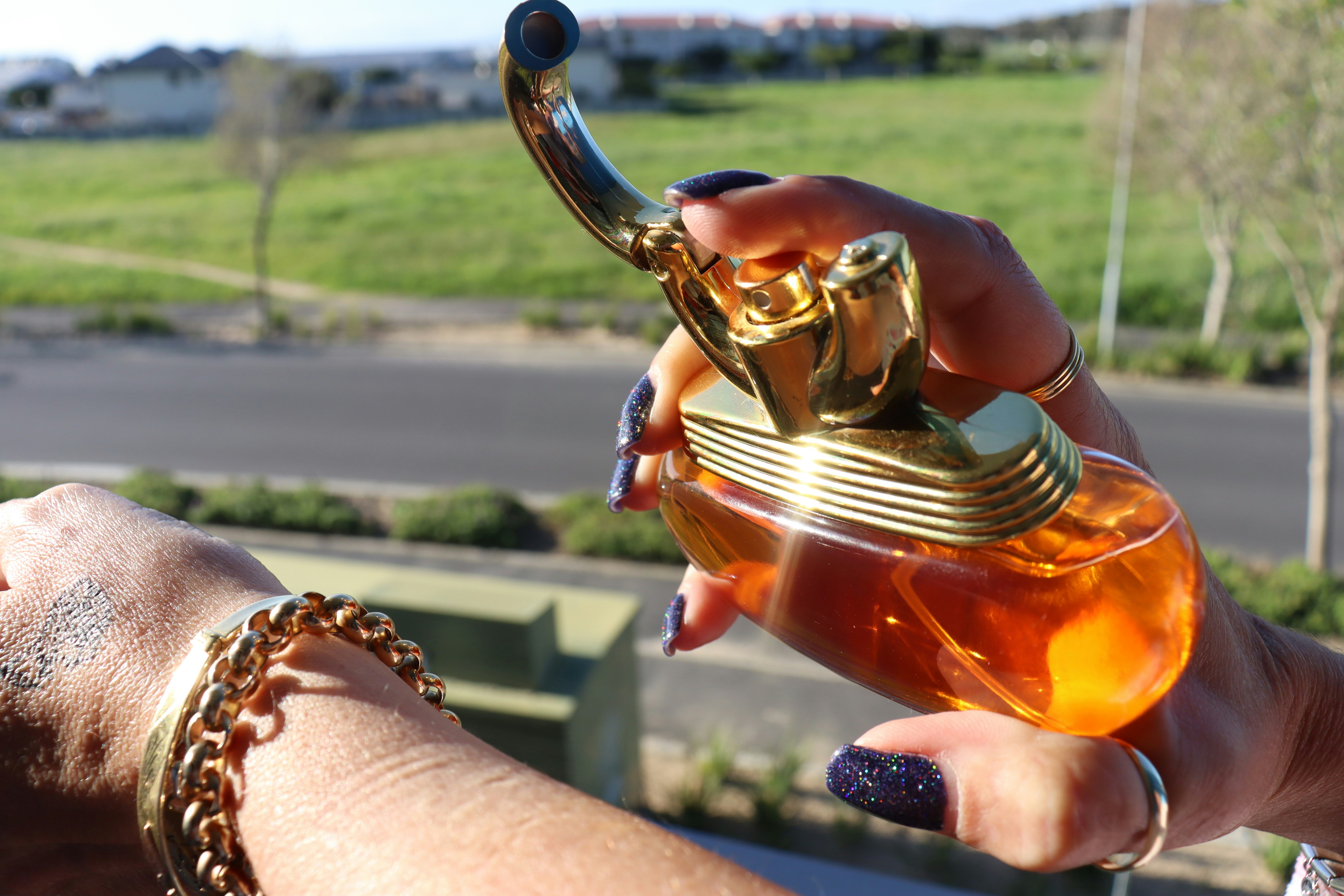 A close-up of a luxurious perfume bottle held by a hand adorned with jewelry, set against a vibrant outdoor backdrop. The golden hues of the bottle reflect sophistication.