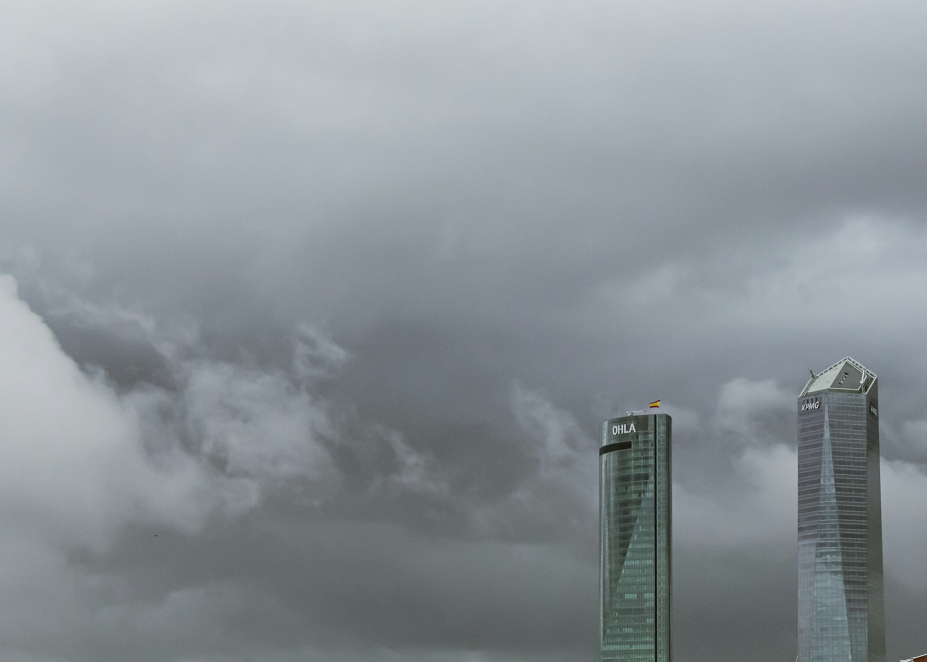 Two modern skyscrapers rise against a backdrop of dark, swirling clouds, evoking a sense of impending weather. The scene captures the stark contrast between architecture and nature.
