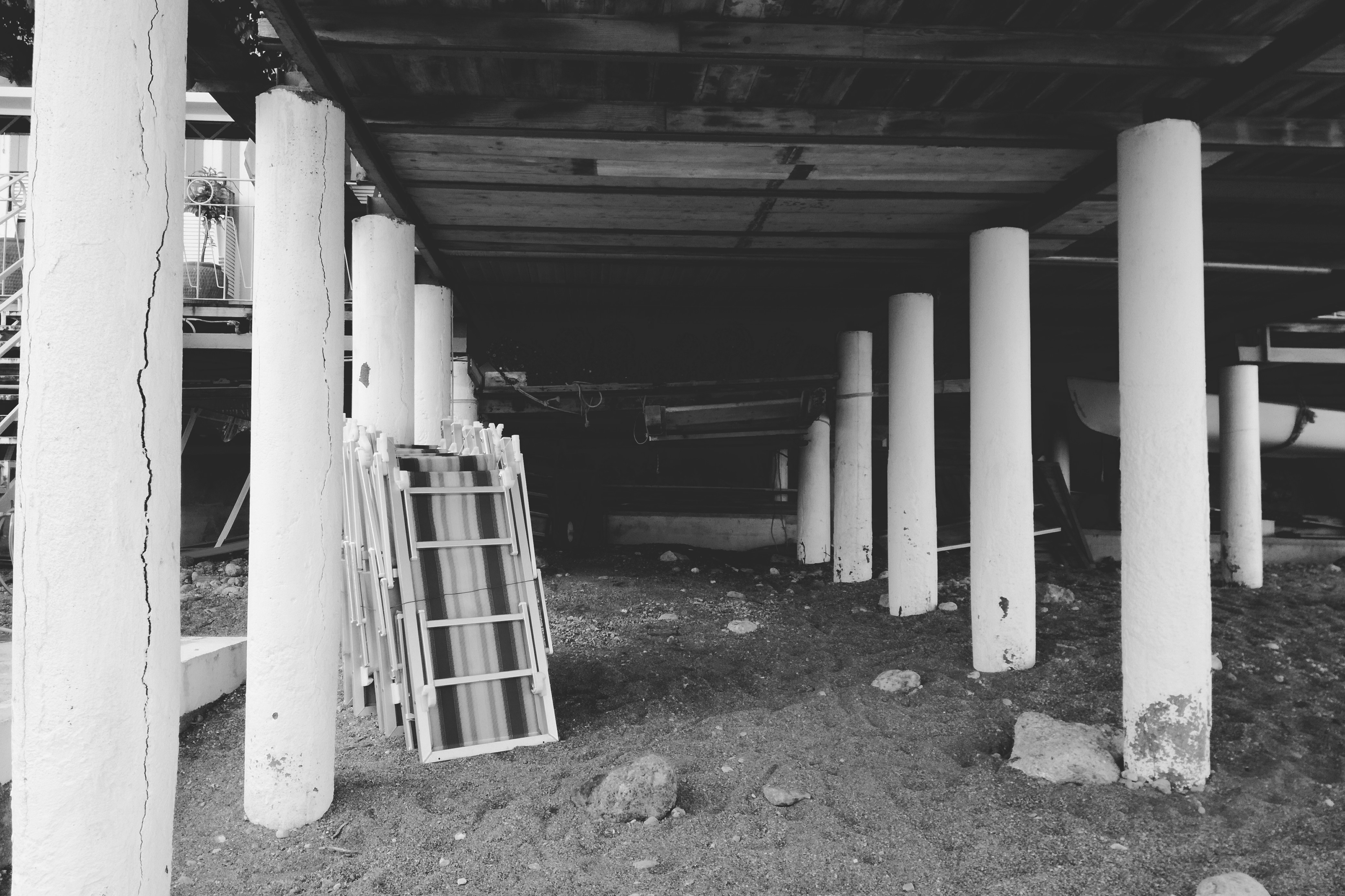 Black-and-white photograph of a shadowy underpass framed by tall concrete columns, with a leaning wooden pallet and scattered rocks guiding the eye toward a distant dark corridor.