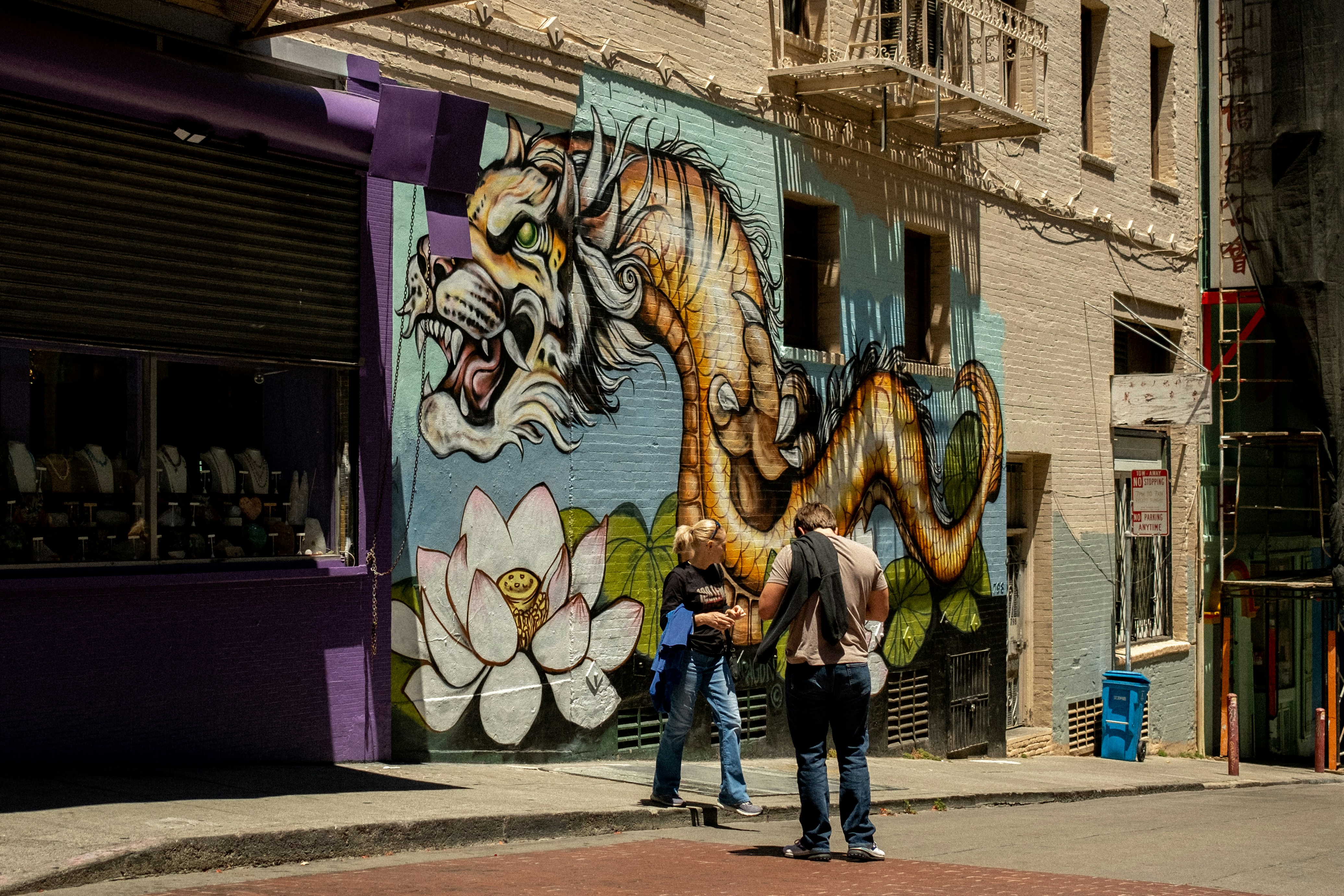 Two visitors discuss beneath a dragon mural in a sunlit urban alley