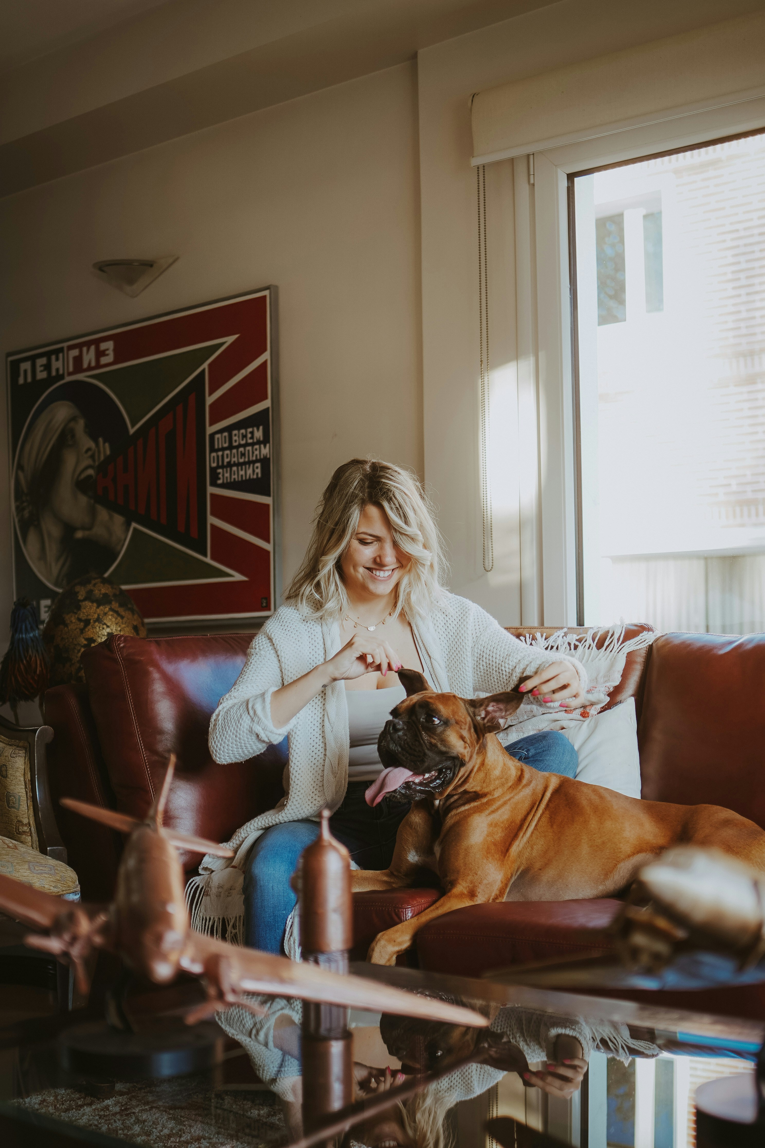 A woman gently brushes her dog while sitting on a cozy couch, surrounded by vintage decor and natural light streaming through the window.