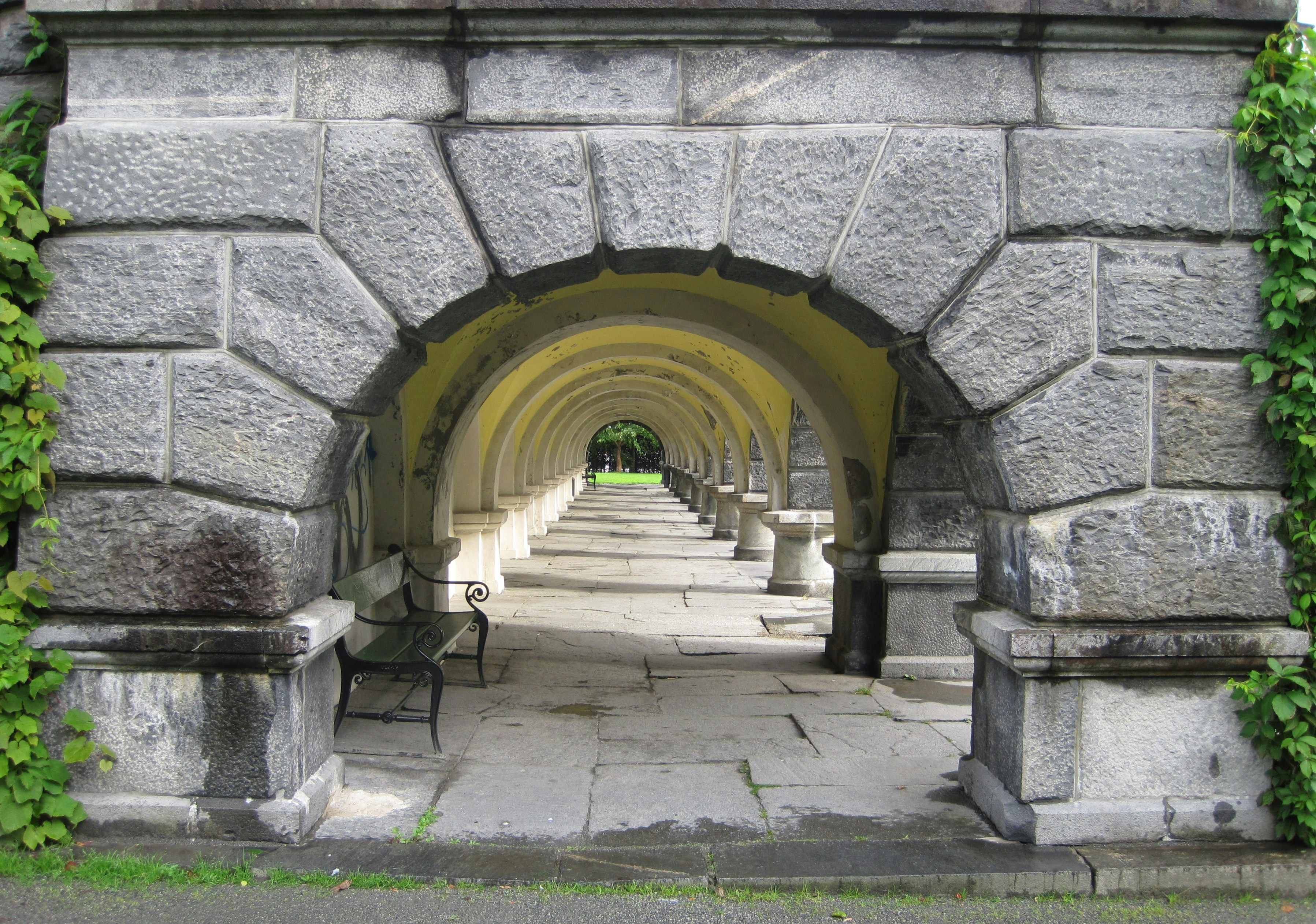 A photograph of a stone archway tunnel along a stone walkway, with ivy-clad sides and a bench.