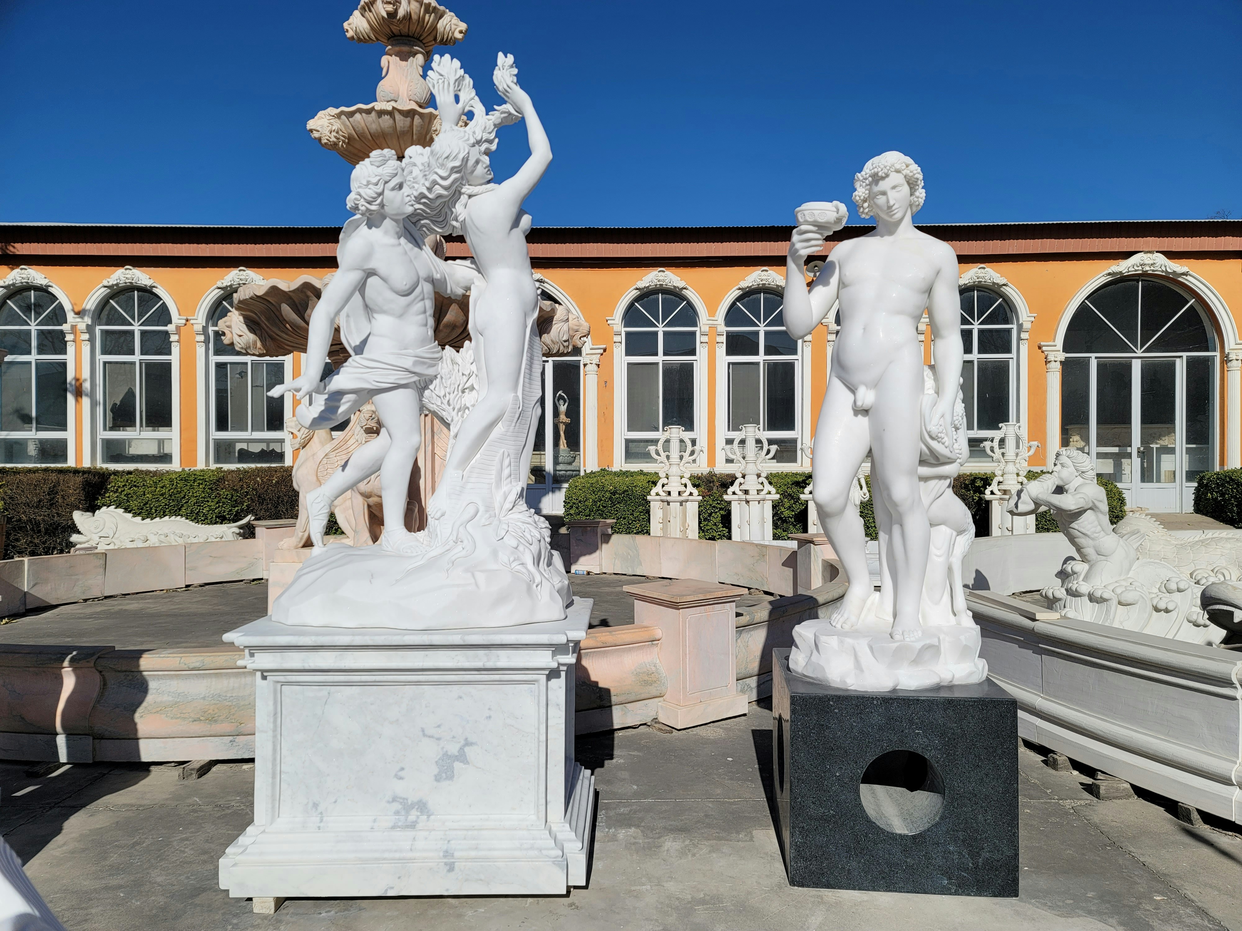 Life-sized marble statues of Apollo and Daphne in a sunlit garden courtyard, with a classical building facade in the background.