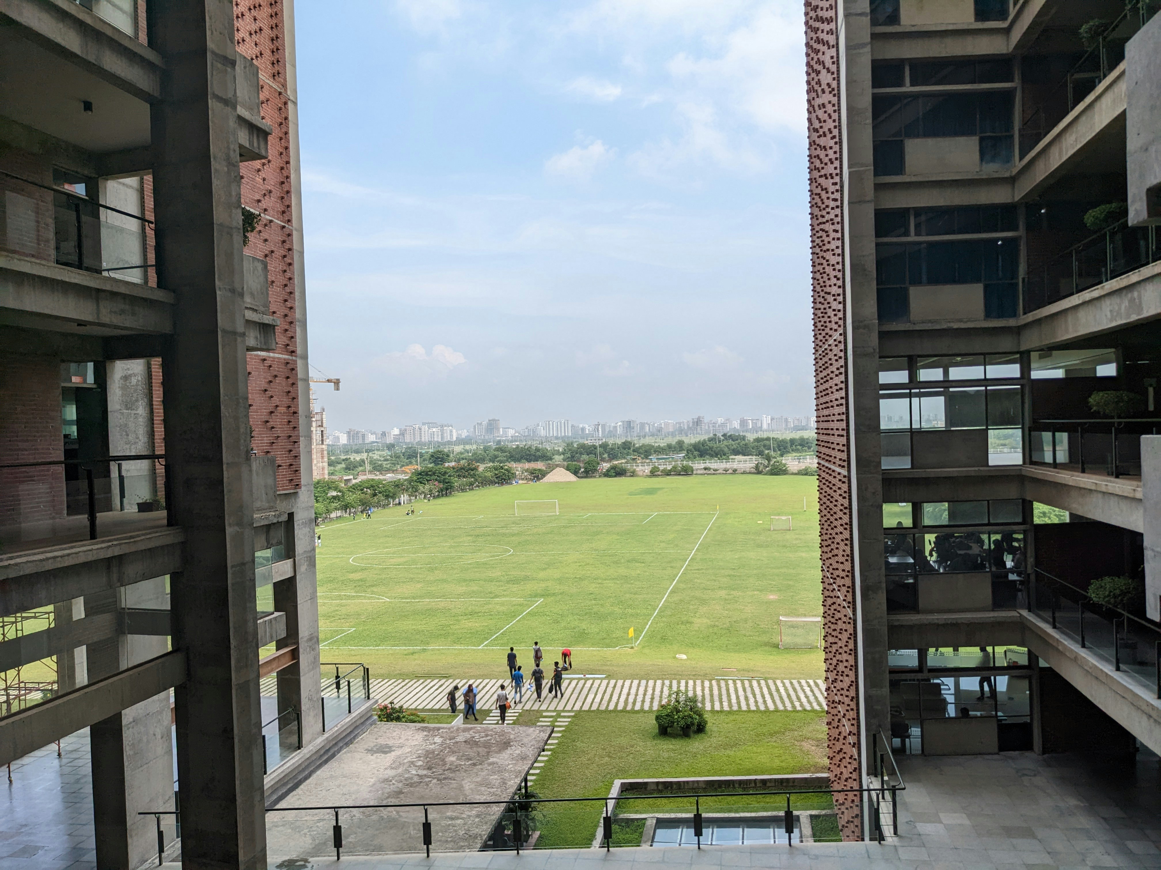 Photograph shows people gathering on a well-kept field framed by two high-rise buildings, with a distant city skyline on the horizon.