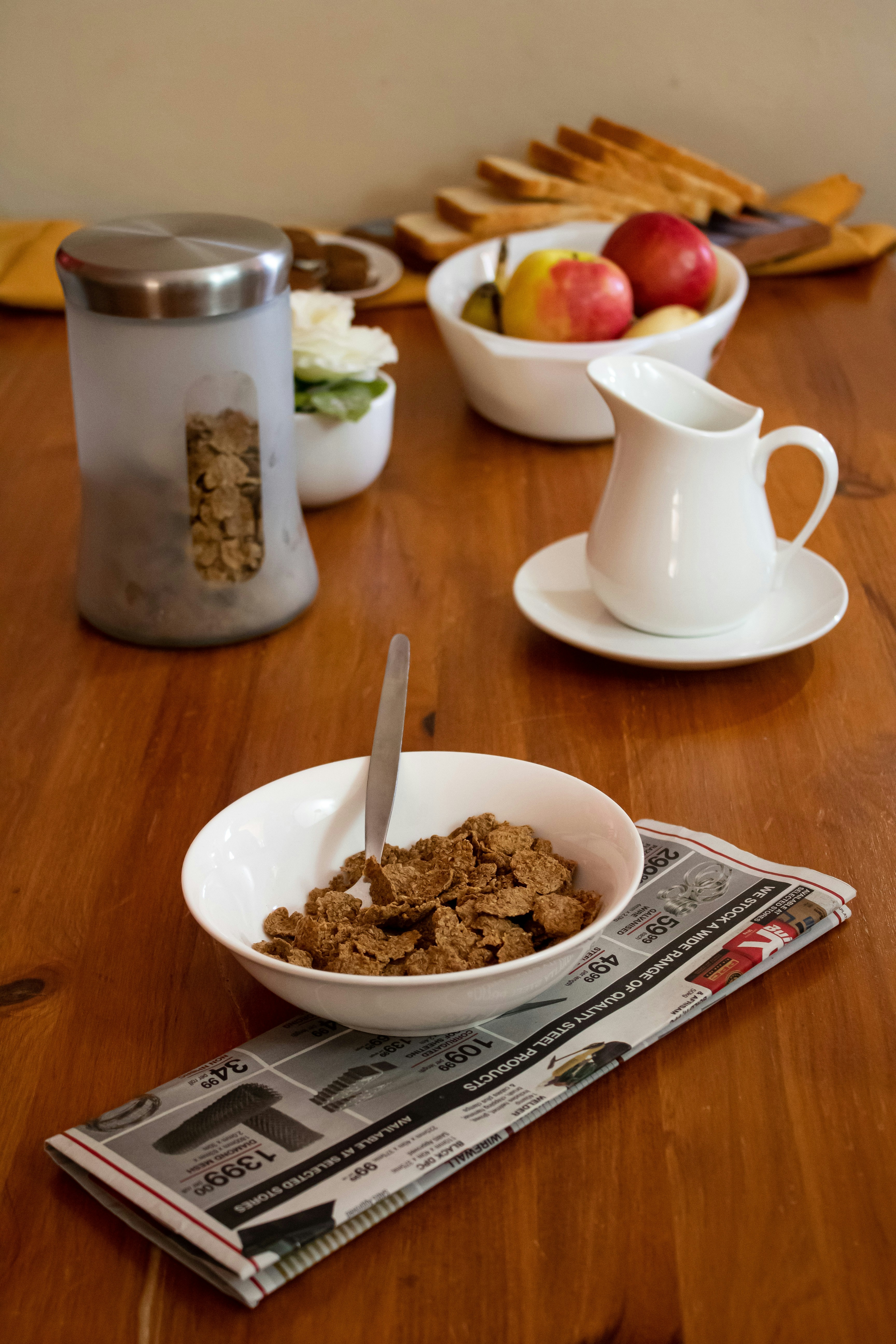 Bowl of cereal on a newspaper placemat surrounded by a milk jug, fruits, and toast on a wooden table.