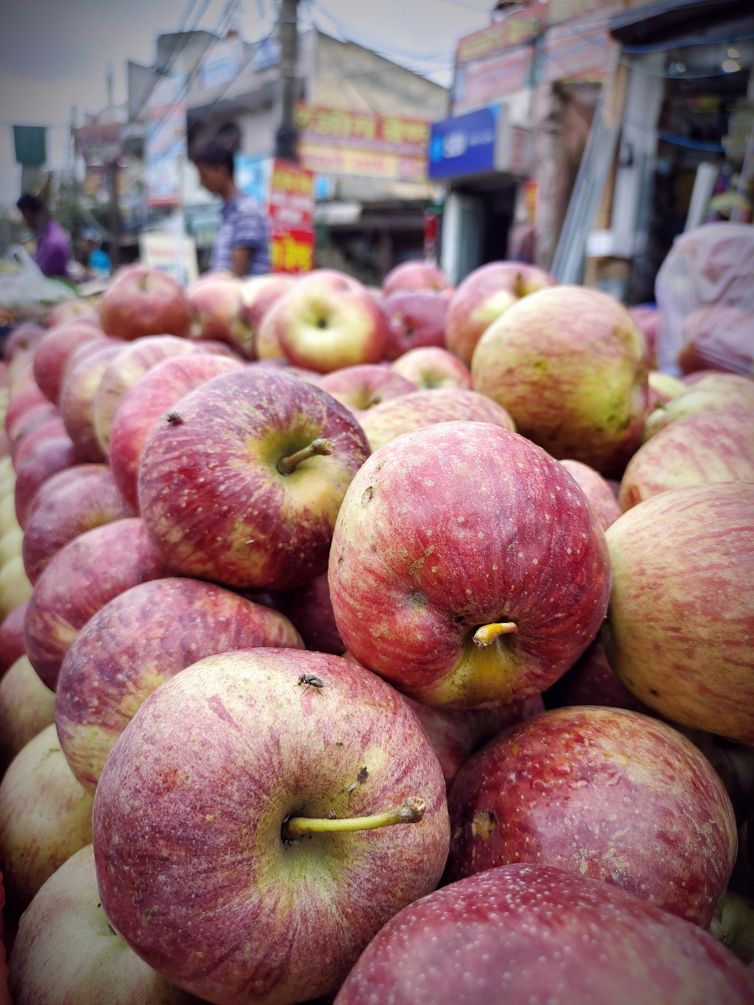 A mound of red apples dominates the foreground while a busy street market blurs in the background, highlighting texture and color. The scene captures fruit presentation amid everyday bustle.
