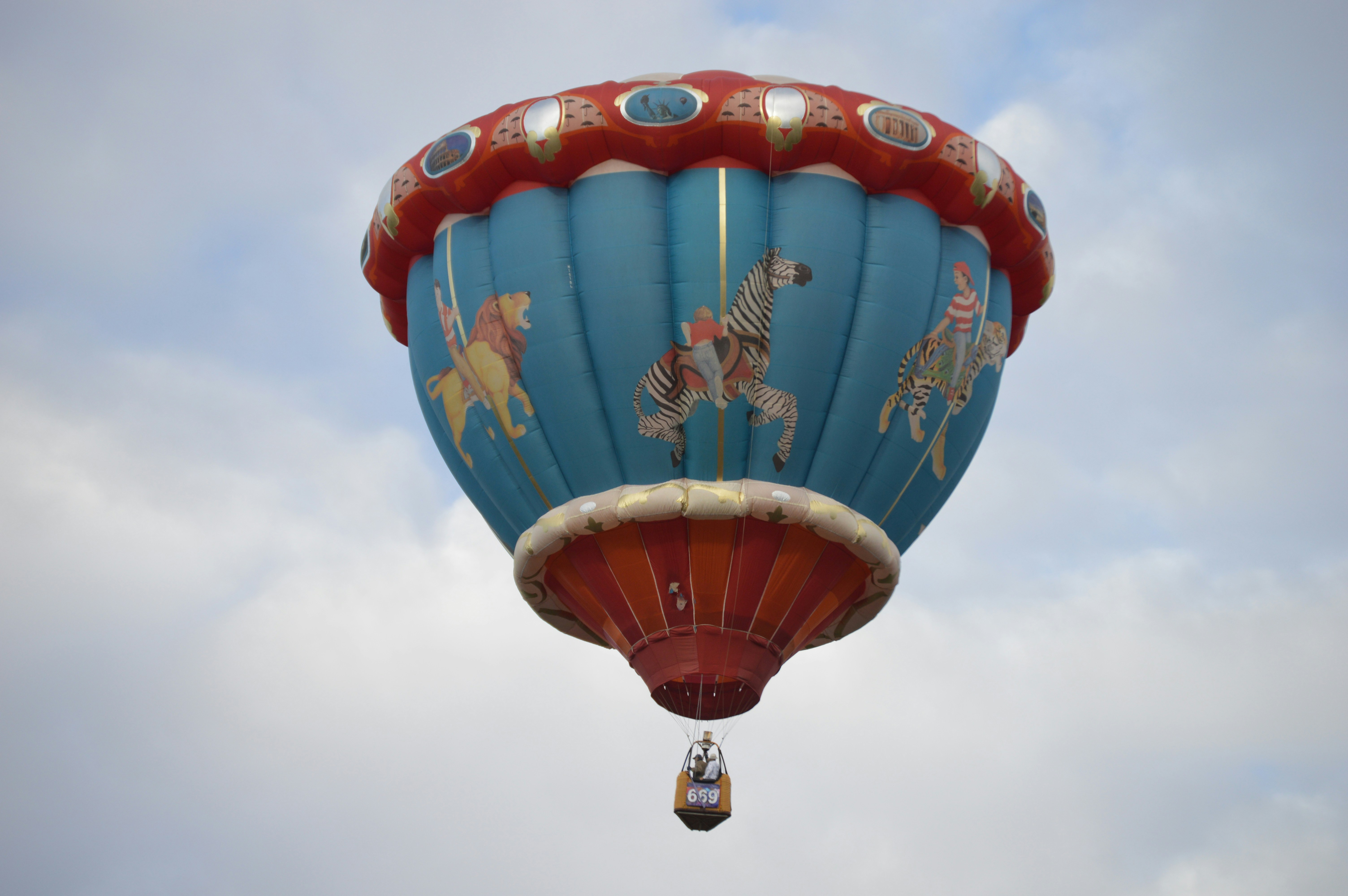 Blue hot air balloon with carousel horse designs soaring against a cloudy sky.