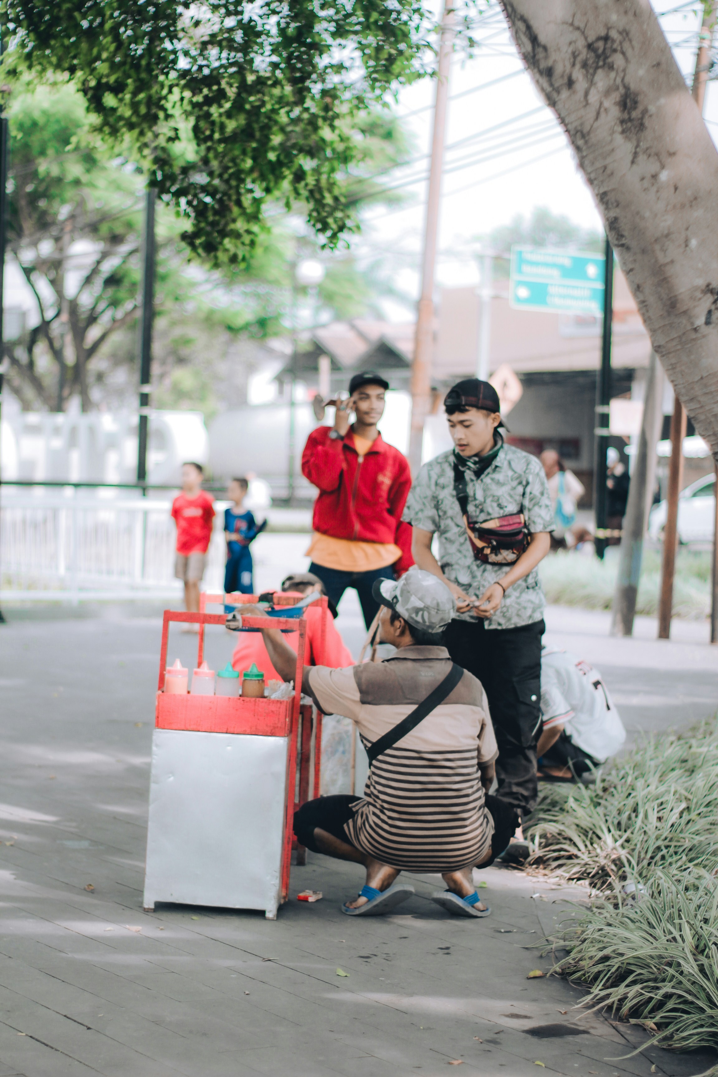 Kneeling Vendor by a Red-White Beverage Cart in a Dappled Street Scene