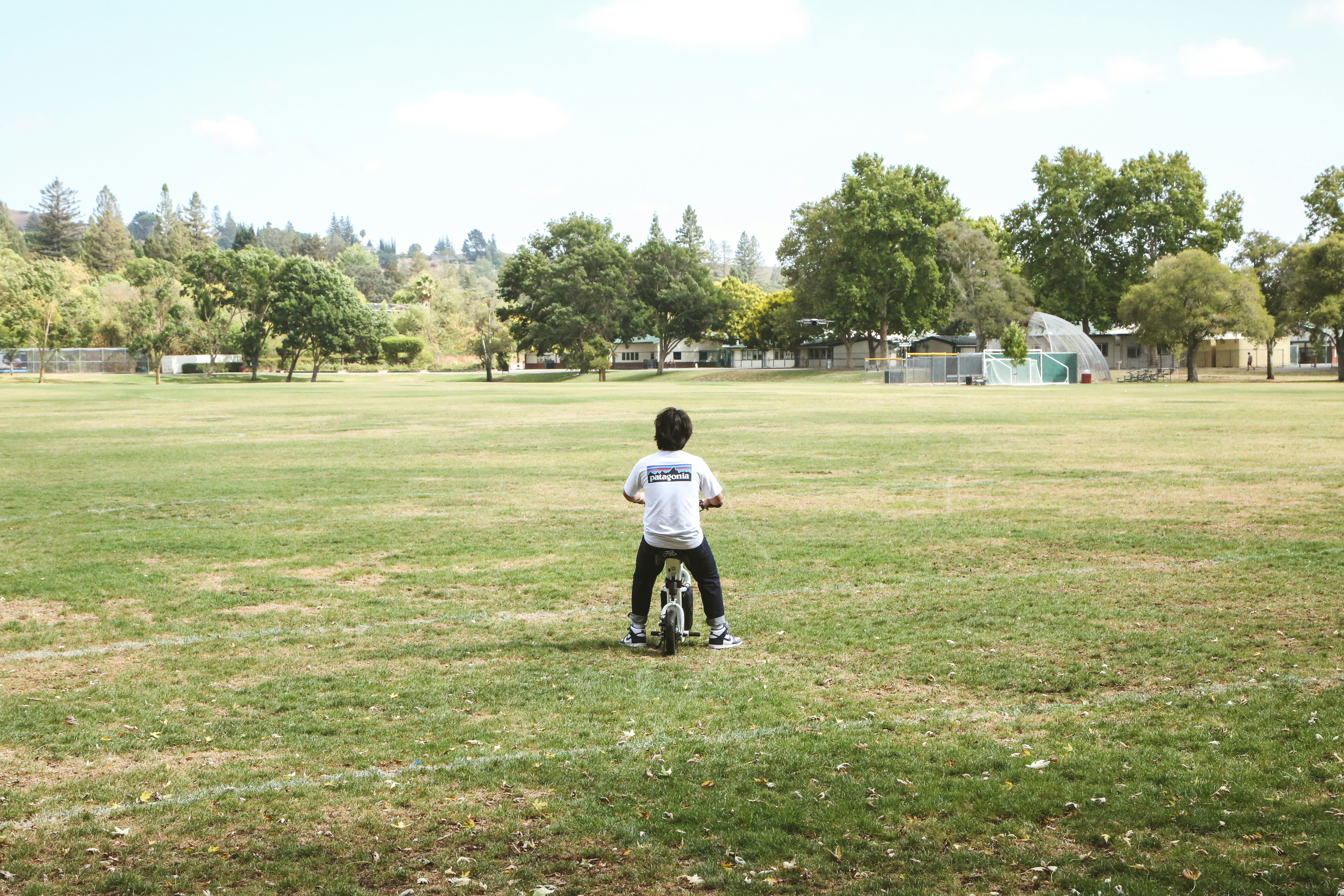 Child riding a bicycle across a wide open green field, surrounded by trees and distant houses under a clear sky.