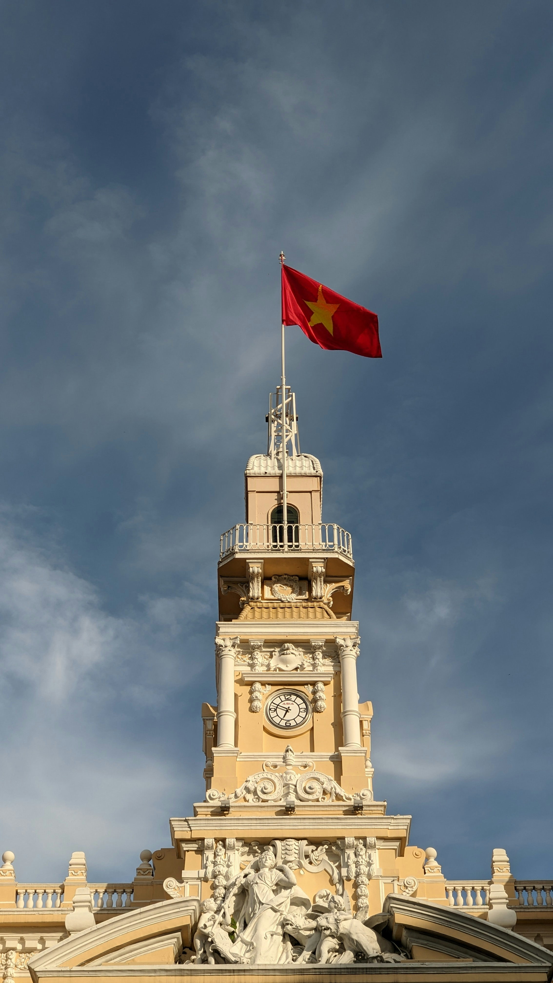 The ornate clock tower of a historic building stands against a backdrop of blue skies, adorned with the Vietnamese flag. A symbol of national pride and architectural elegance.