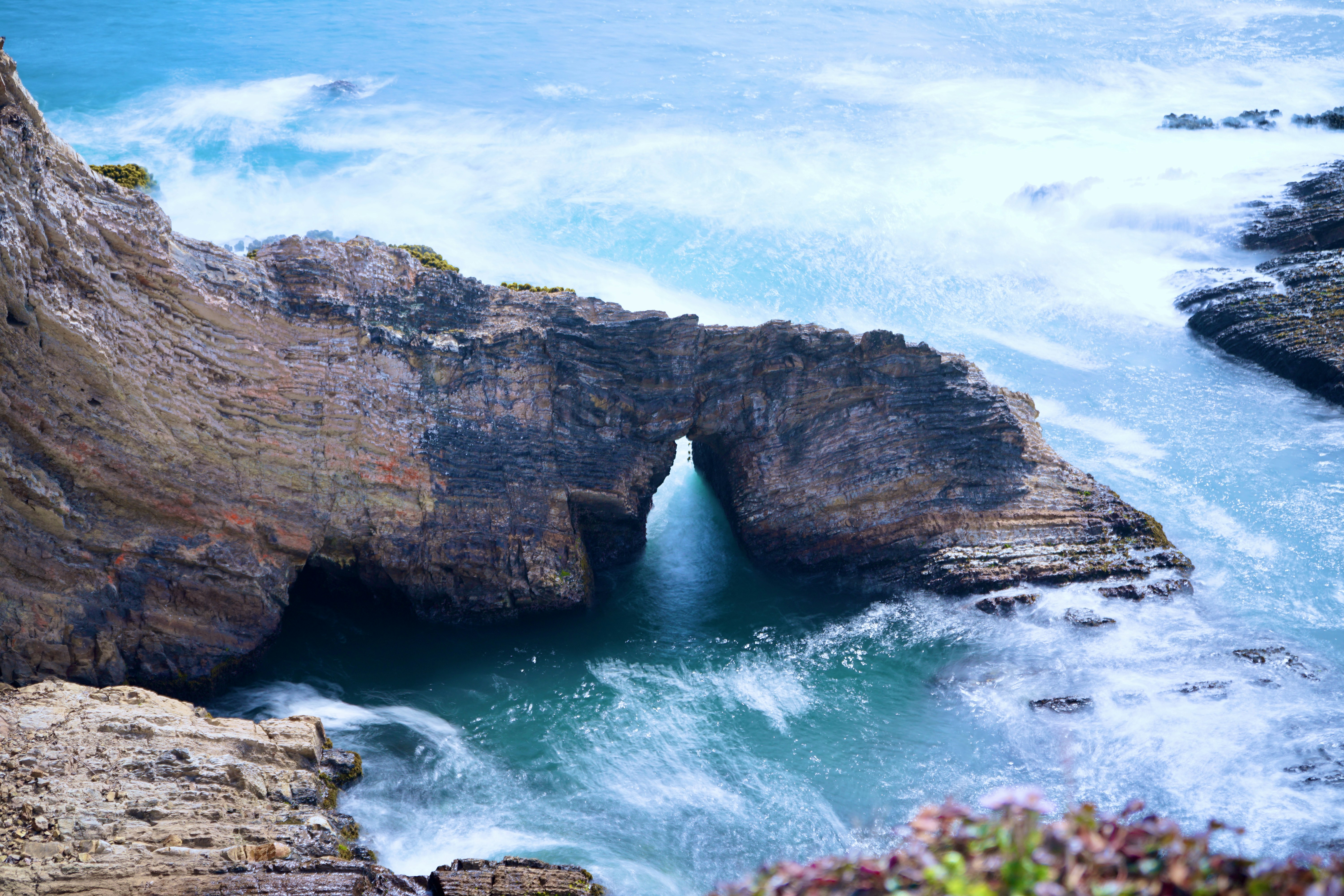 Natural rock archway with waves crashing through, surrounded by rugged cliffs and vibrant blue water.