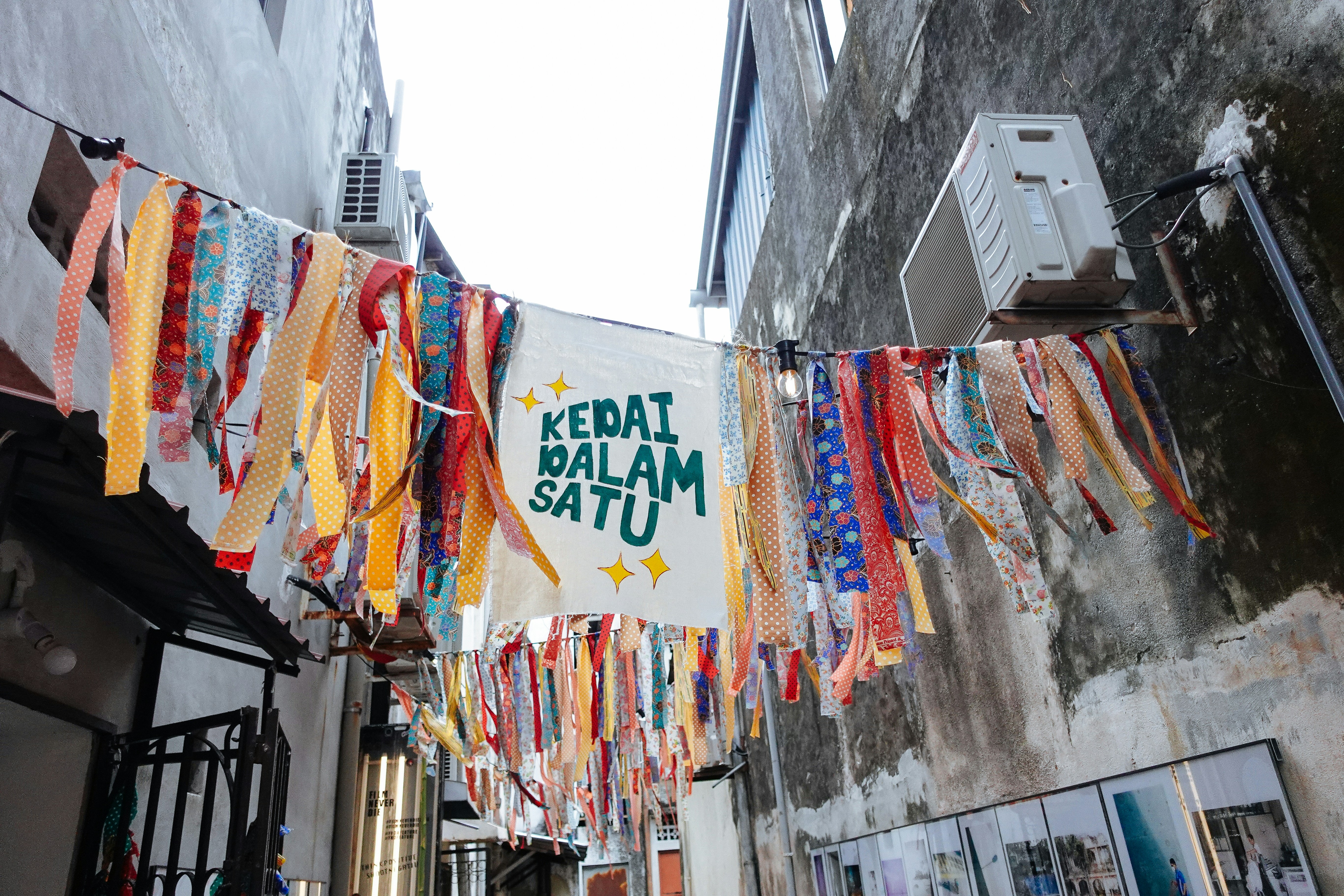 Vibrant fabric strips hang between buildings in a narrow alley, adorned with a sign that reads 'KEDAI DALAM SATU.'