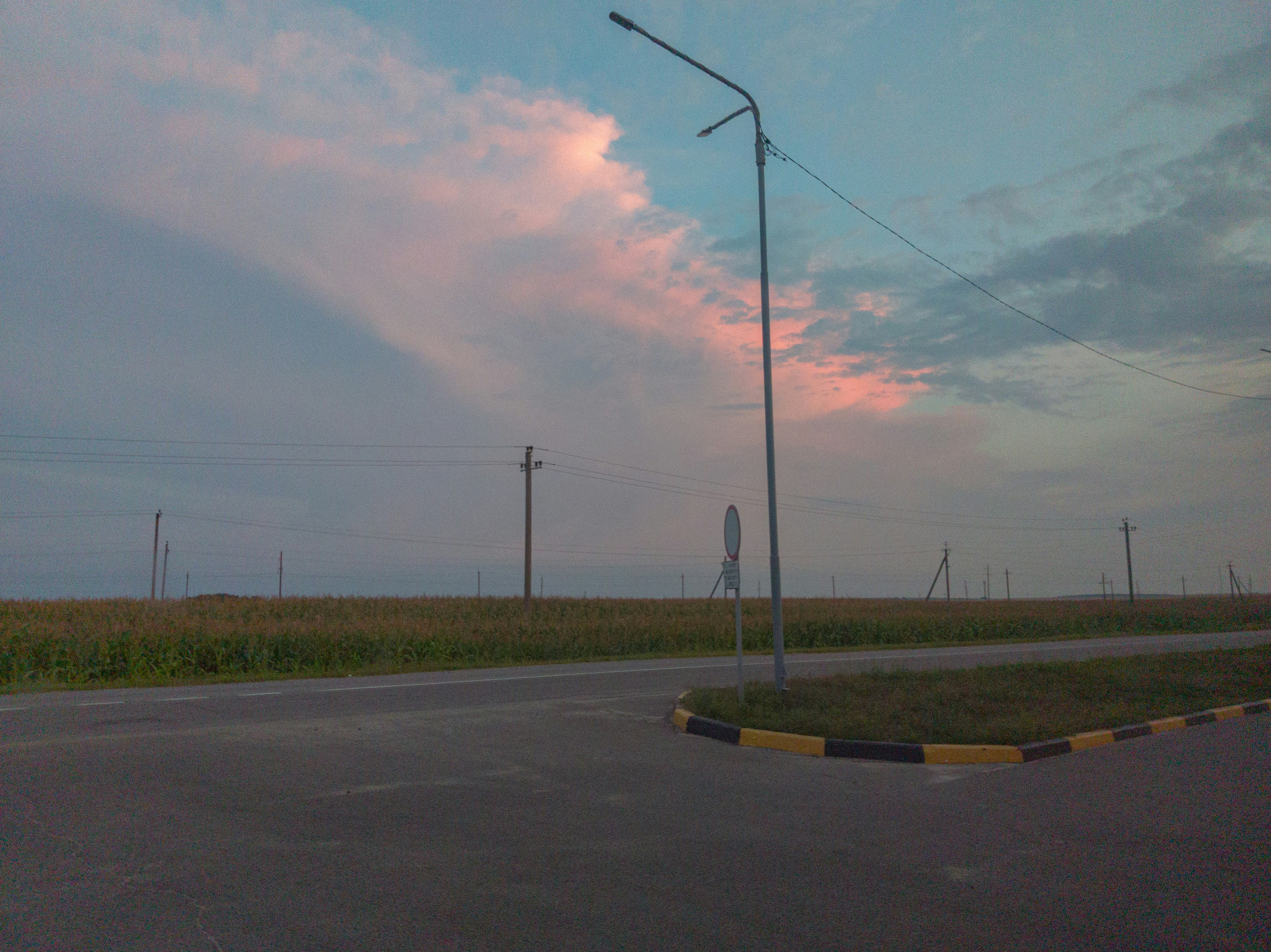 Soft pastel sky over a rural road at dusk with a lone street lamp, power poles, and a yellow-black curb.