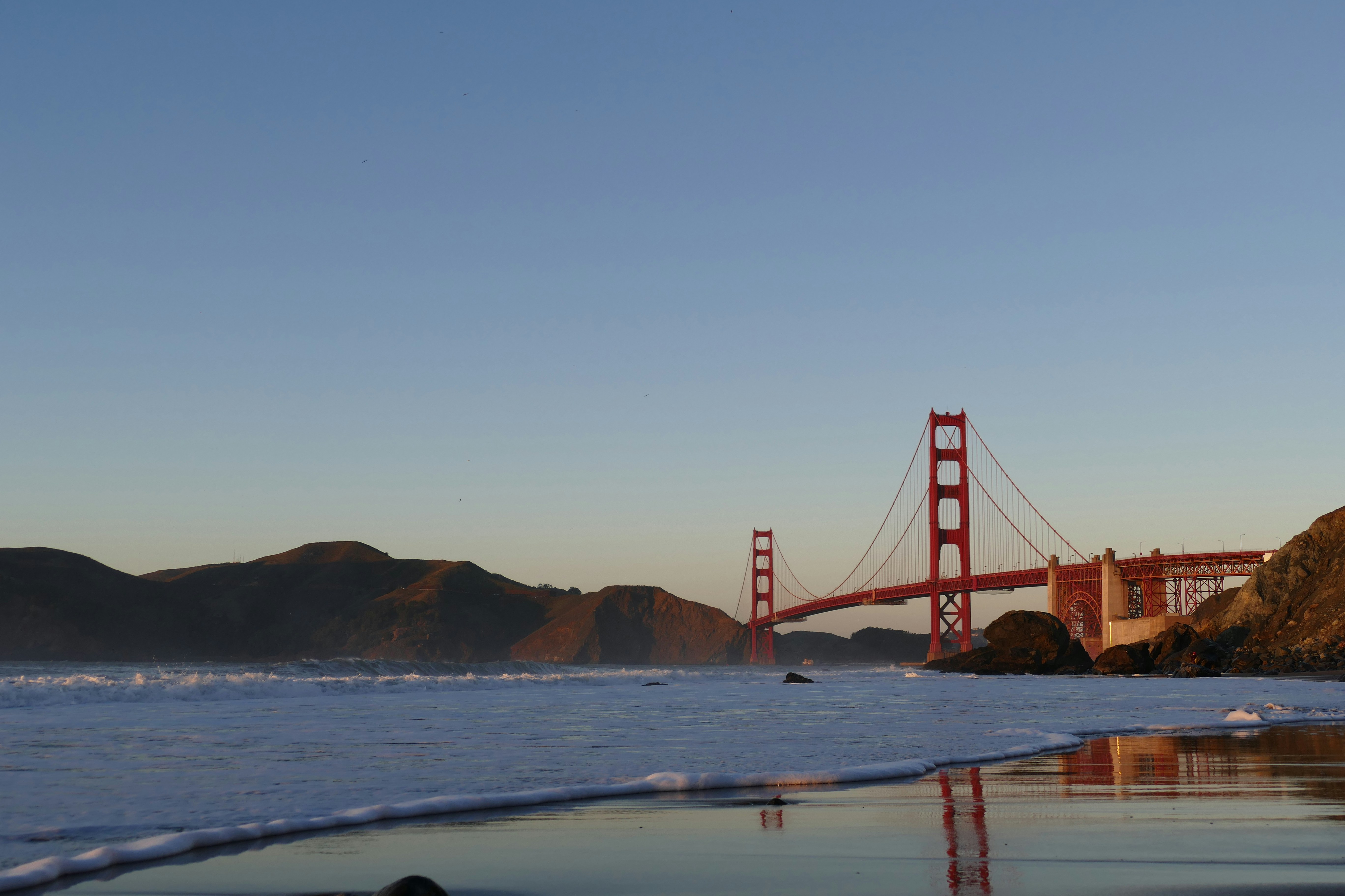 Golden Gate Bridge silhouetted against a clear sky, reflecting in the calm waters of the beach at sunset.