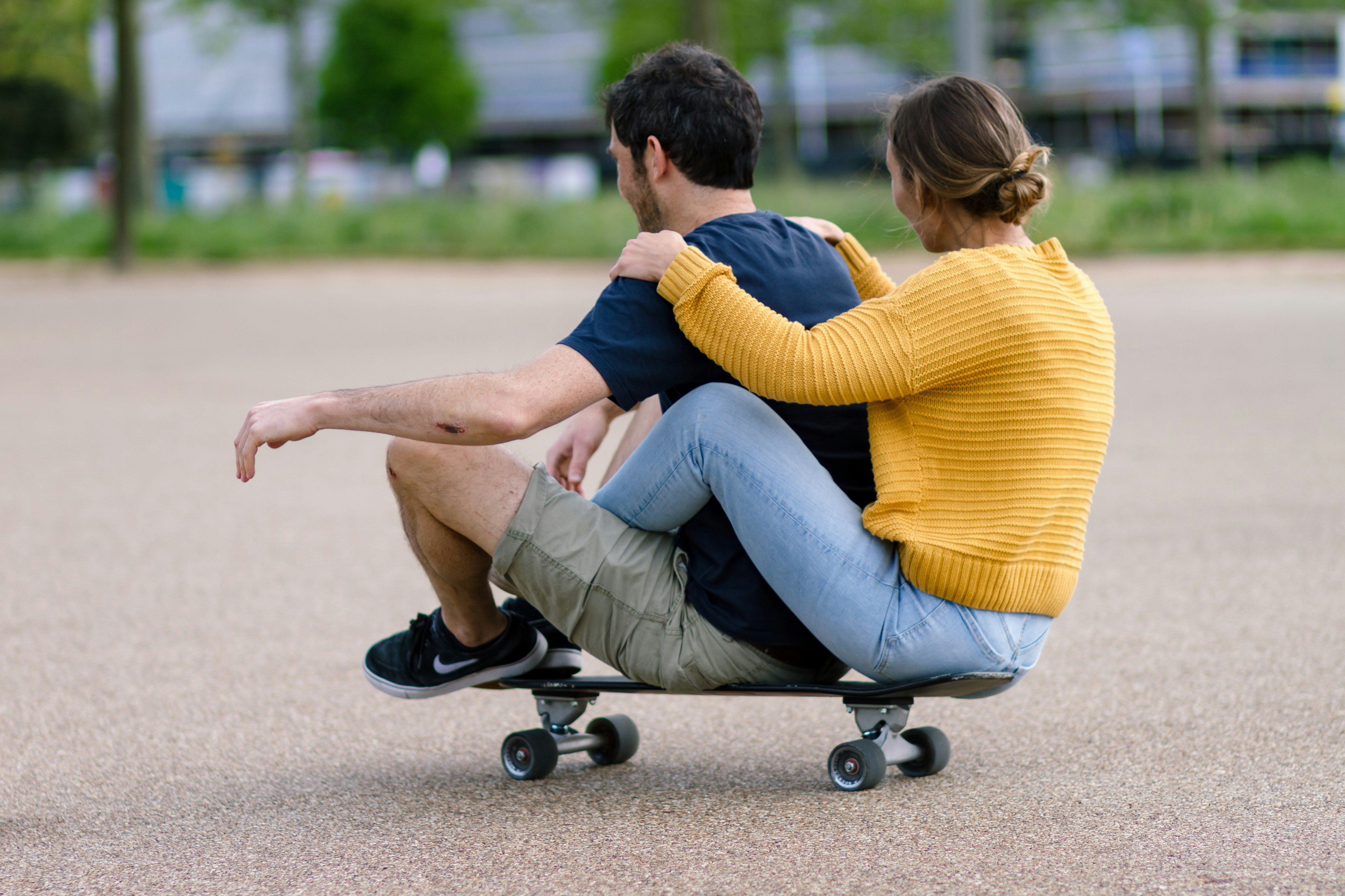 Two people sitting back-to-back on a skateboard gliding smoothly on a paved path.