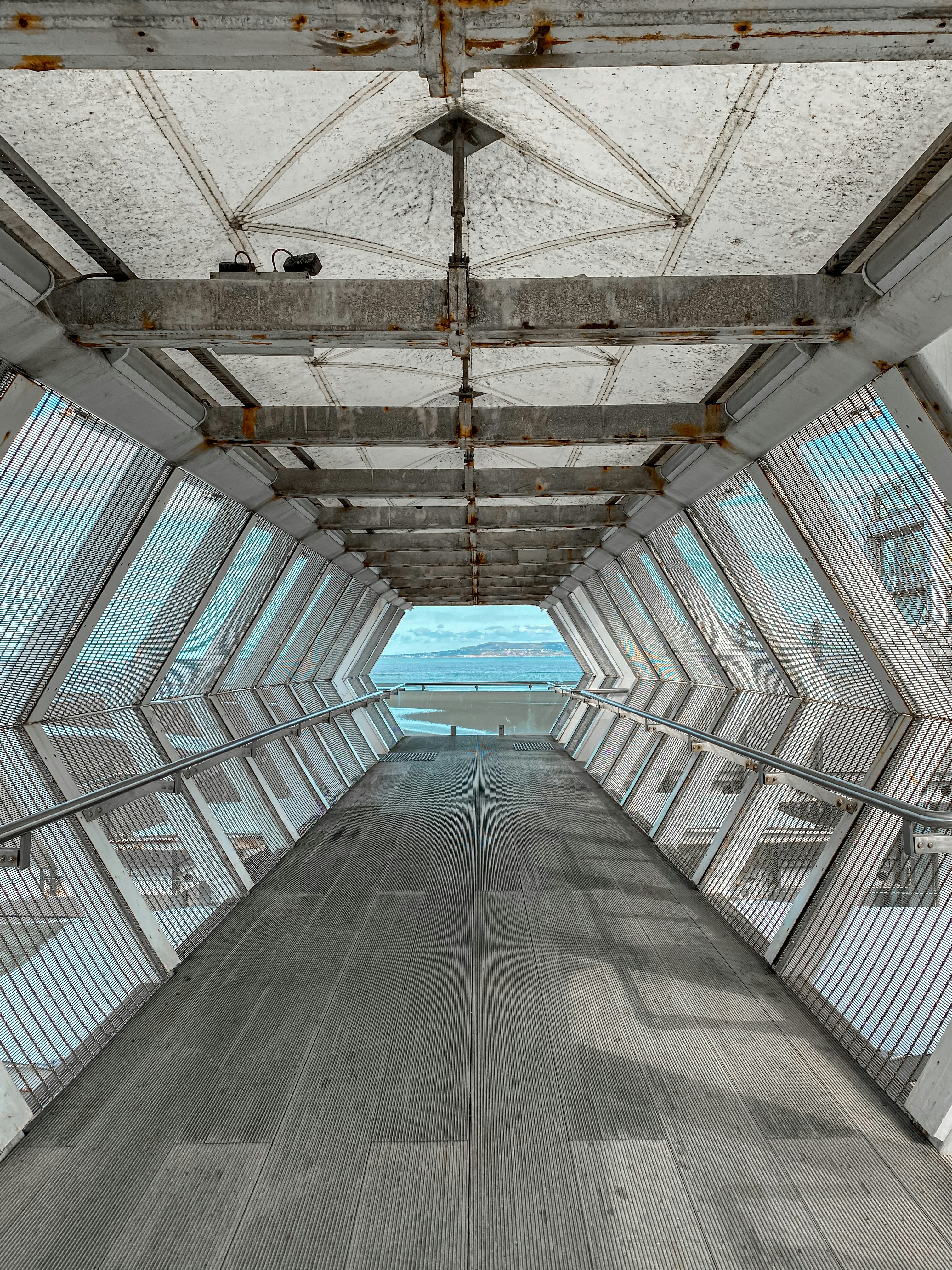 A modern architectural walkway featuring a series of angular glass panels and metal structures, leading to a distant view of the horizon.