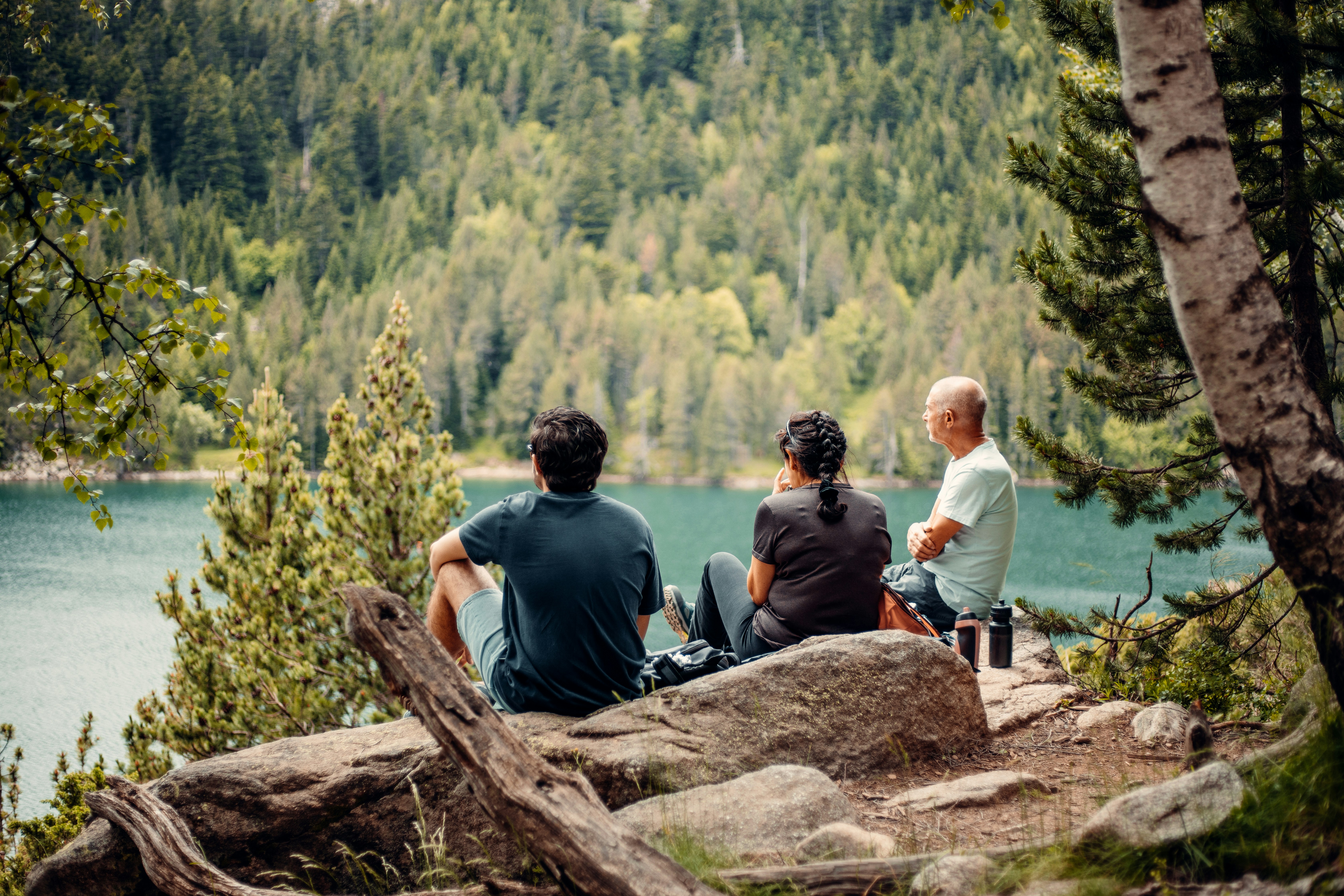 Three people sitting on rocks, gazing at a serene lake surrounded by lush forest.