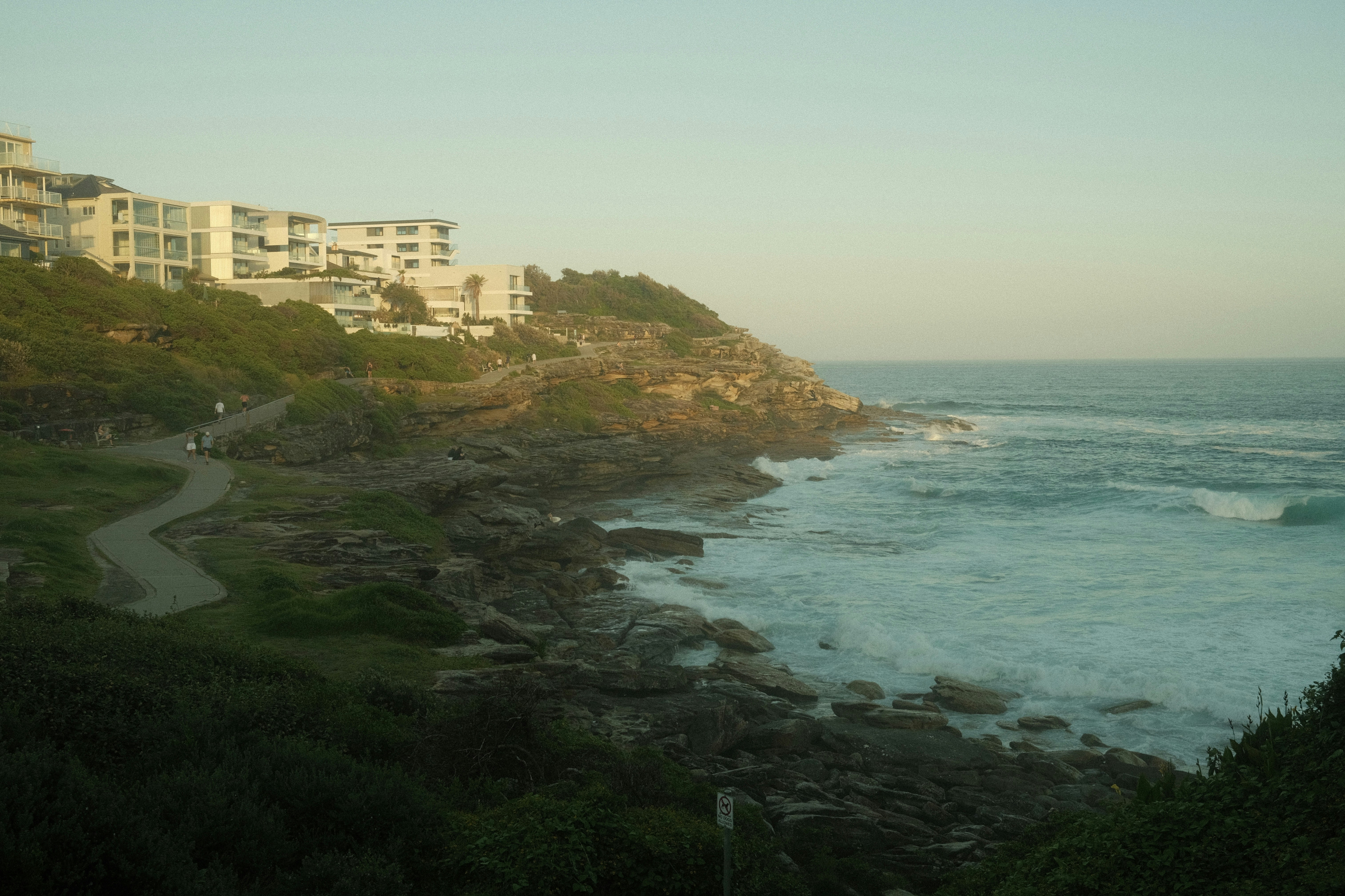 Coastal path winding past modern buildings with waves crashing on rocky shores under a soft morning light.