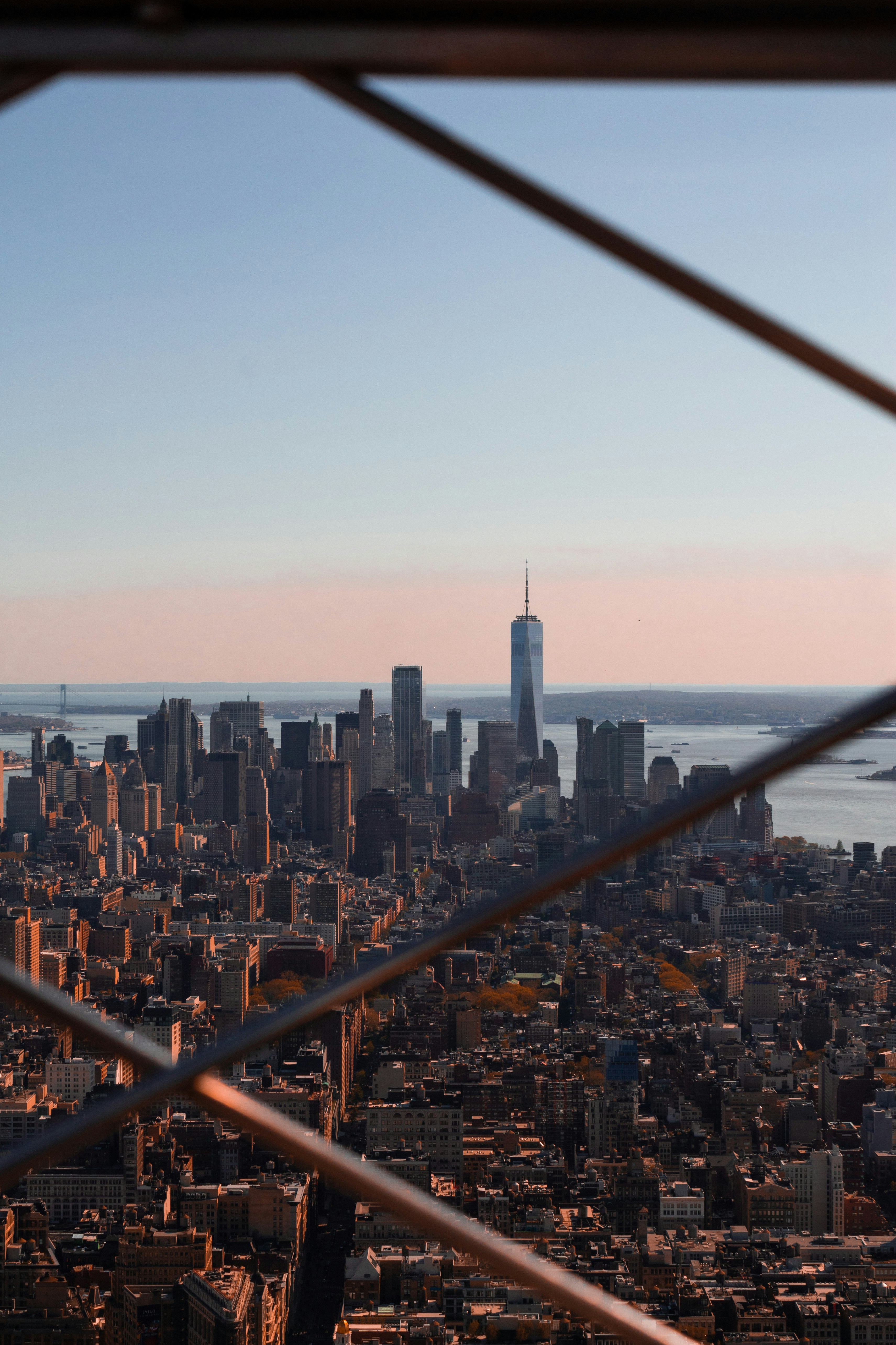 A sweeping view of New York City skyline featuring One World Trade Center, framed by intersecting metal beams. The scene captures the vibrant urban landscape at dusk.