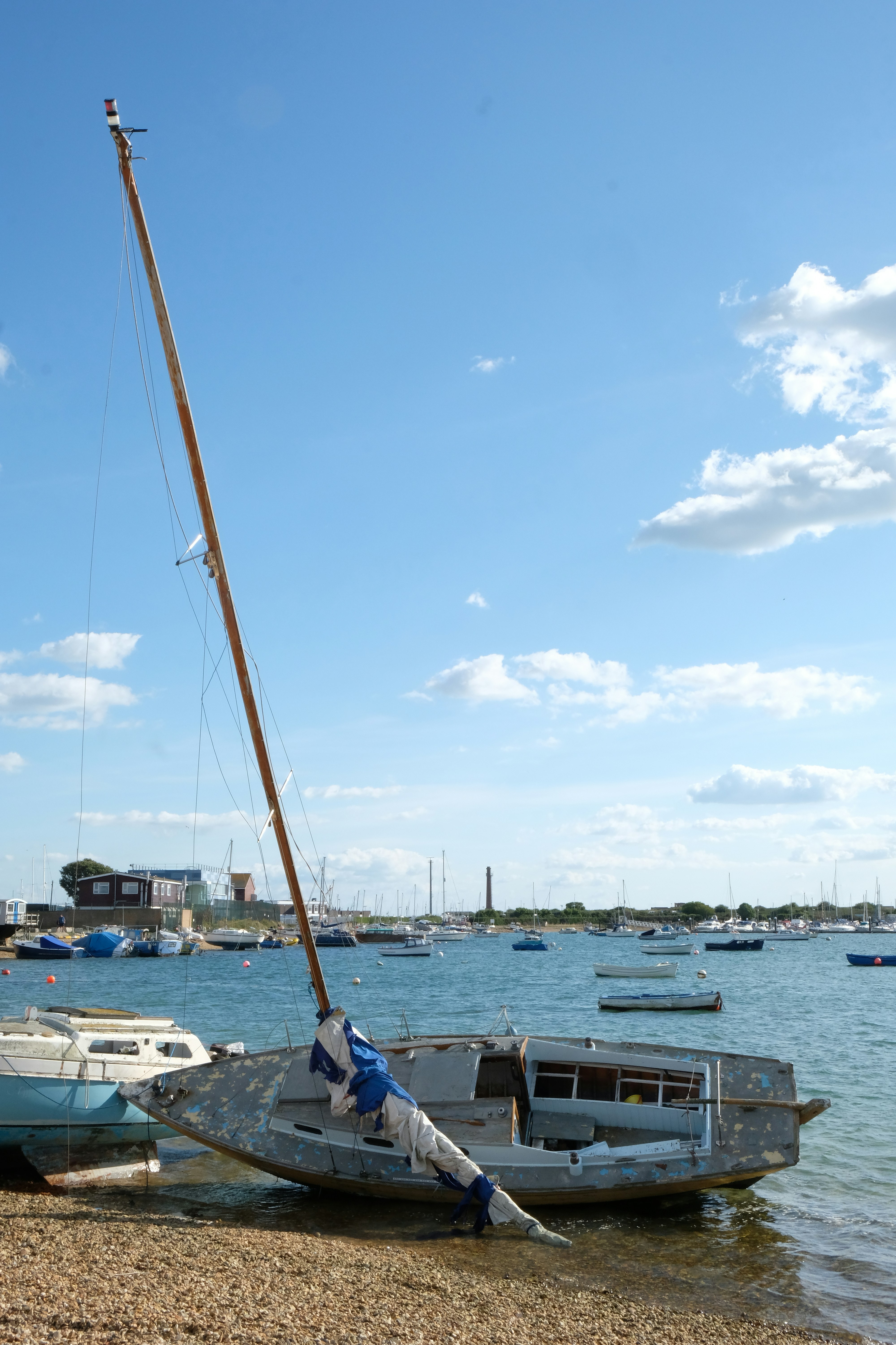 A weathered sailboat rests on a pebbled shore, with a backdrop of a serene harbor dotted with moored vessels under a bright blue sky.