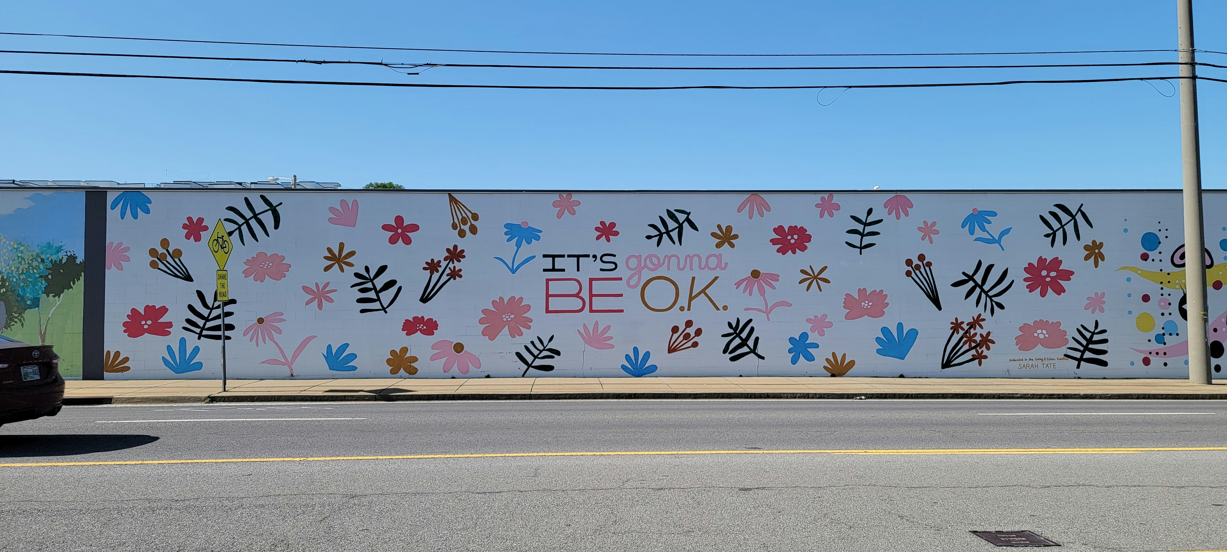 Colorful mural with flowers and the phrase 'It's Going to Be OK' on a long building wall under a clear blue sky.