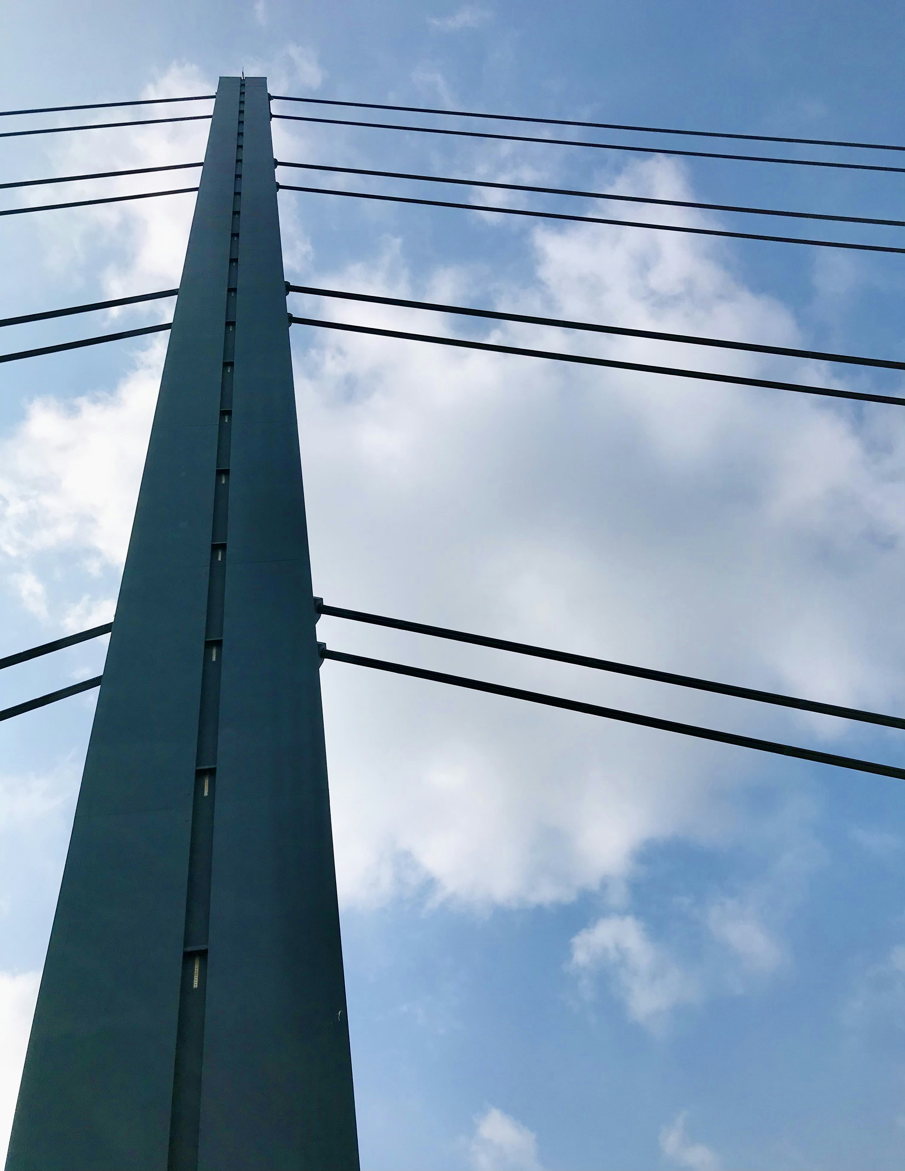 A towering bridge support rises against a backdrop of blue sky and fluffy clouds, showcasing modern engineering. The cables stretch elegantly, emphasizing the structure's height and design.