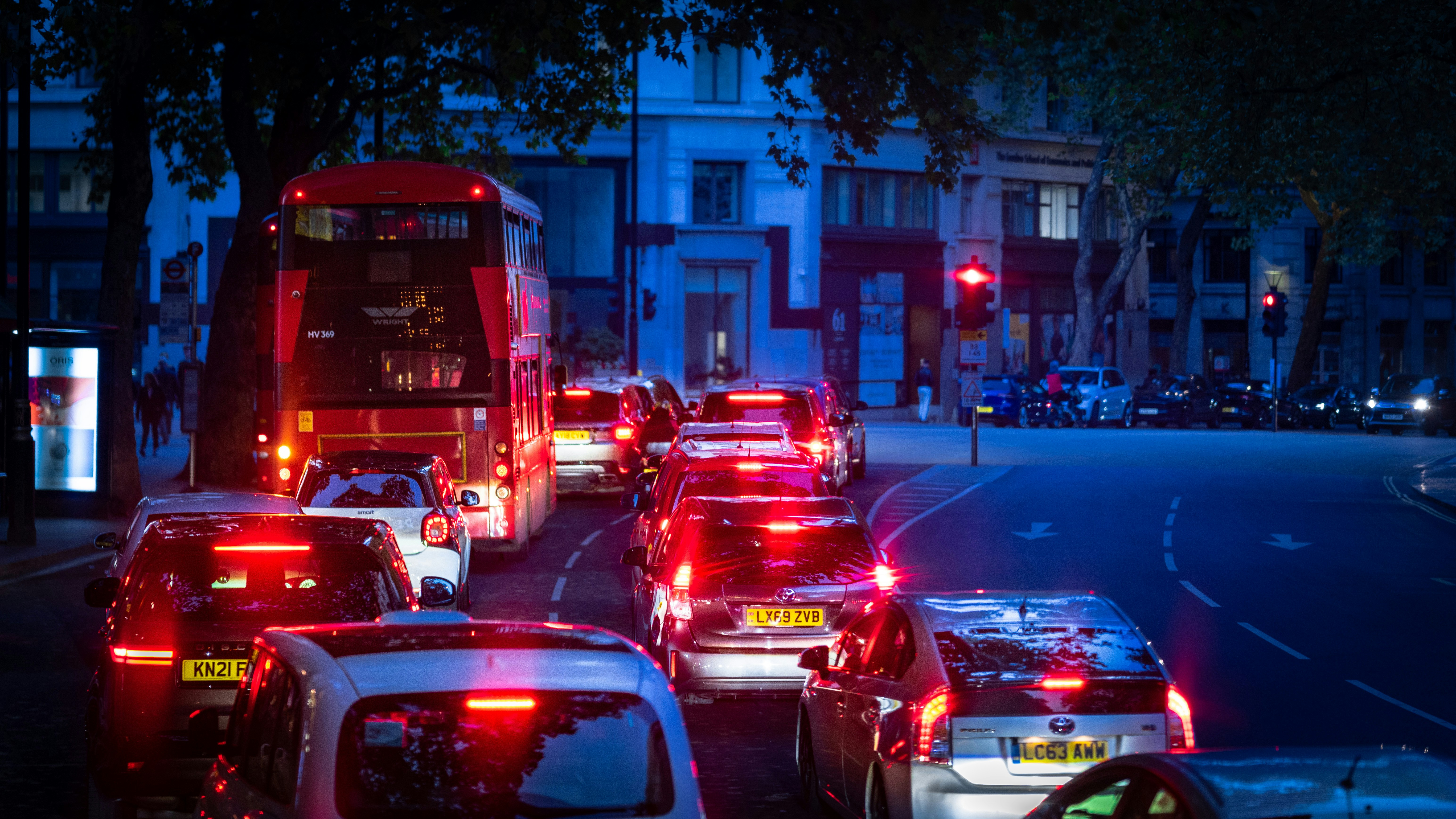 Nighttime city street bustling with traffic, featuring a red double-decker bus and various vehicles illuminated by streetlights and brake lights.