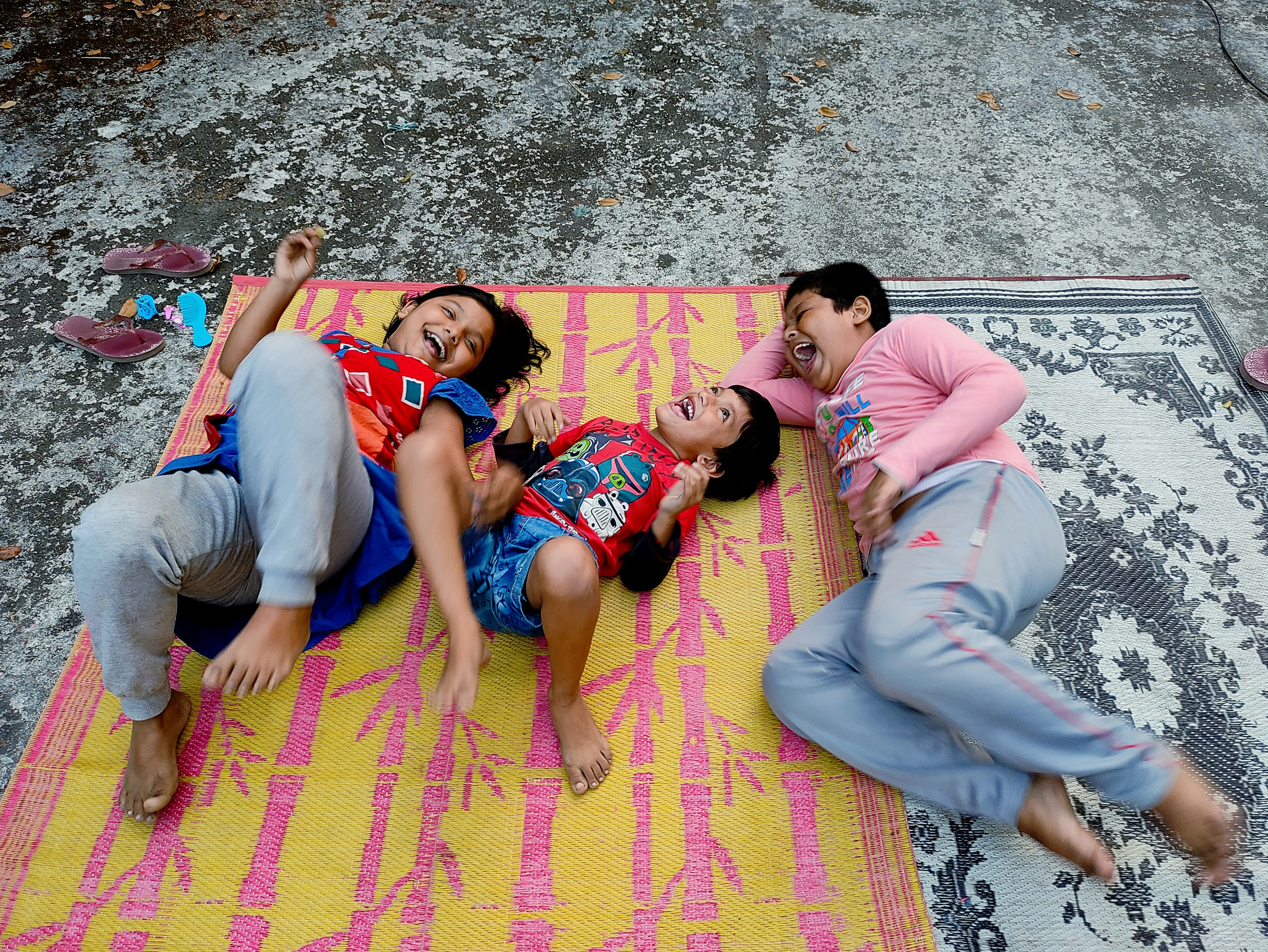 Three people lying on a colorful mat, sharing a moment of laughter on a concrete rooftop.