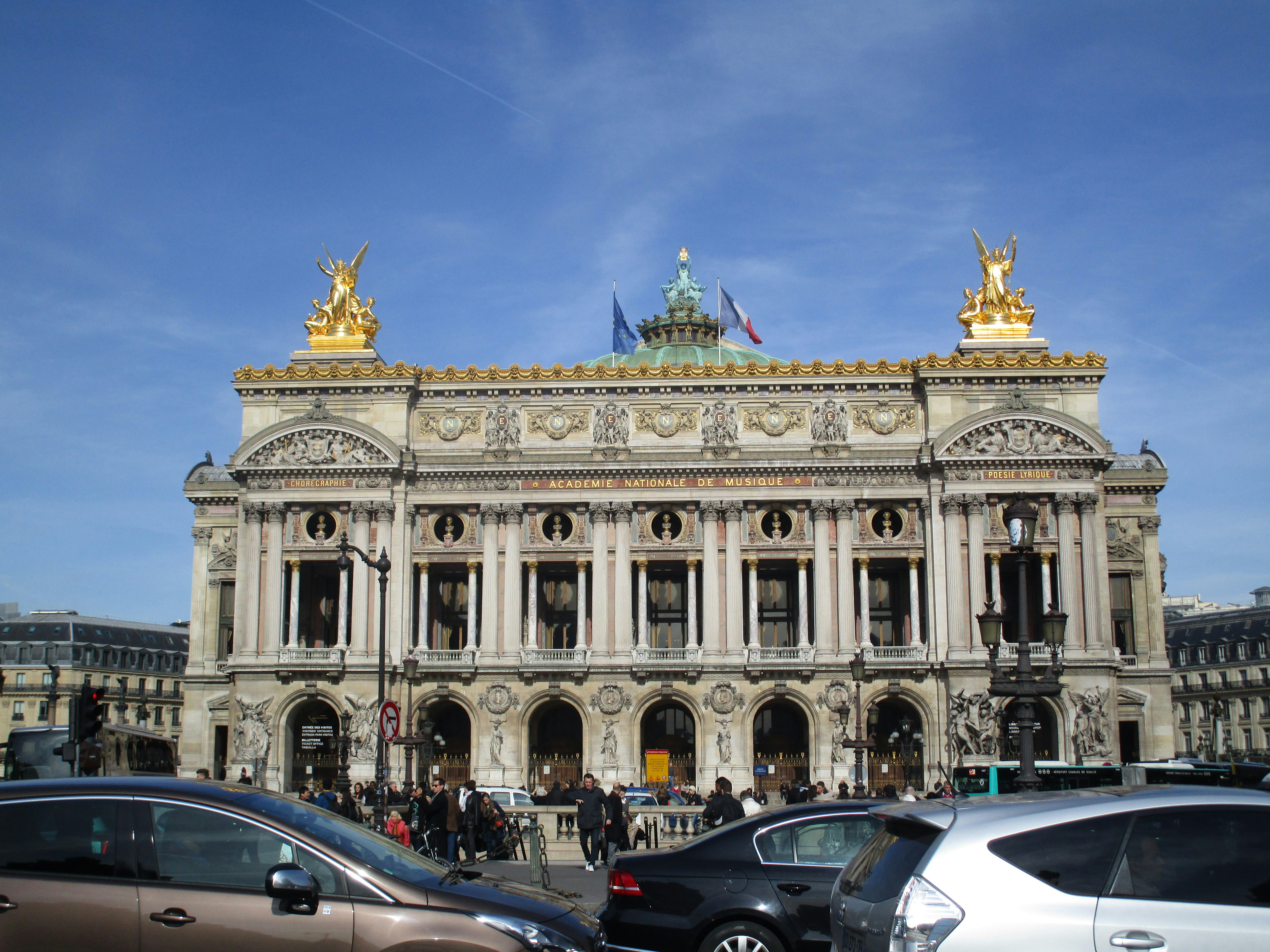 Paris Opéra Garnier's ornate marble facade, flanked by gilded statues, with a busy foreground of parked cars.