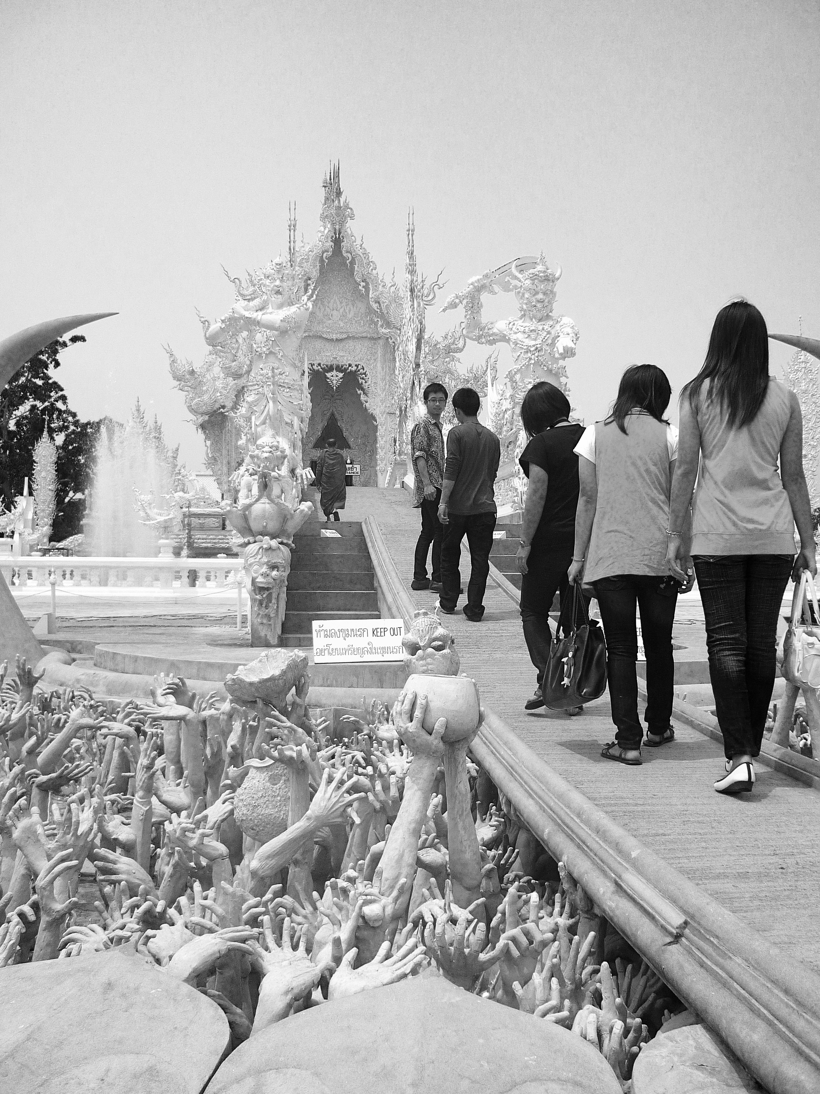Visitors traverse a bridge leading to a stunning white temple, surrounded by intricate sculptures of hands reaching upward. The scene evokes a sense of exploration and spirituality.