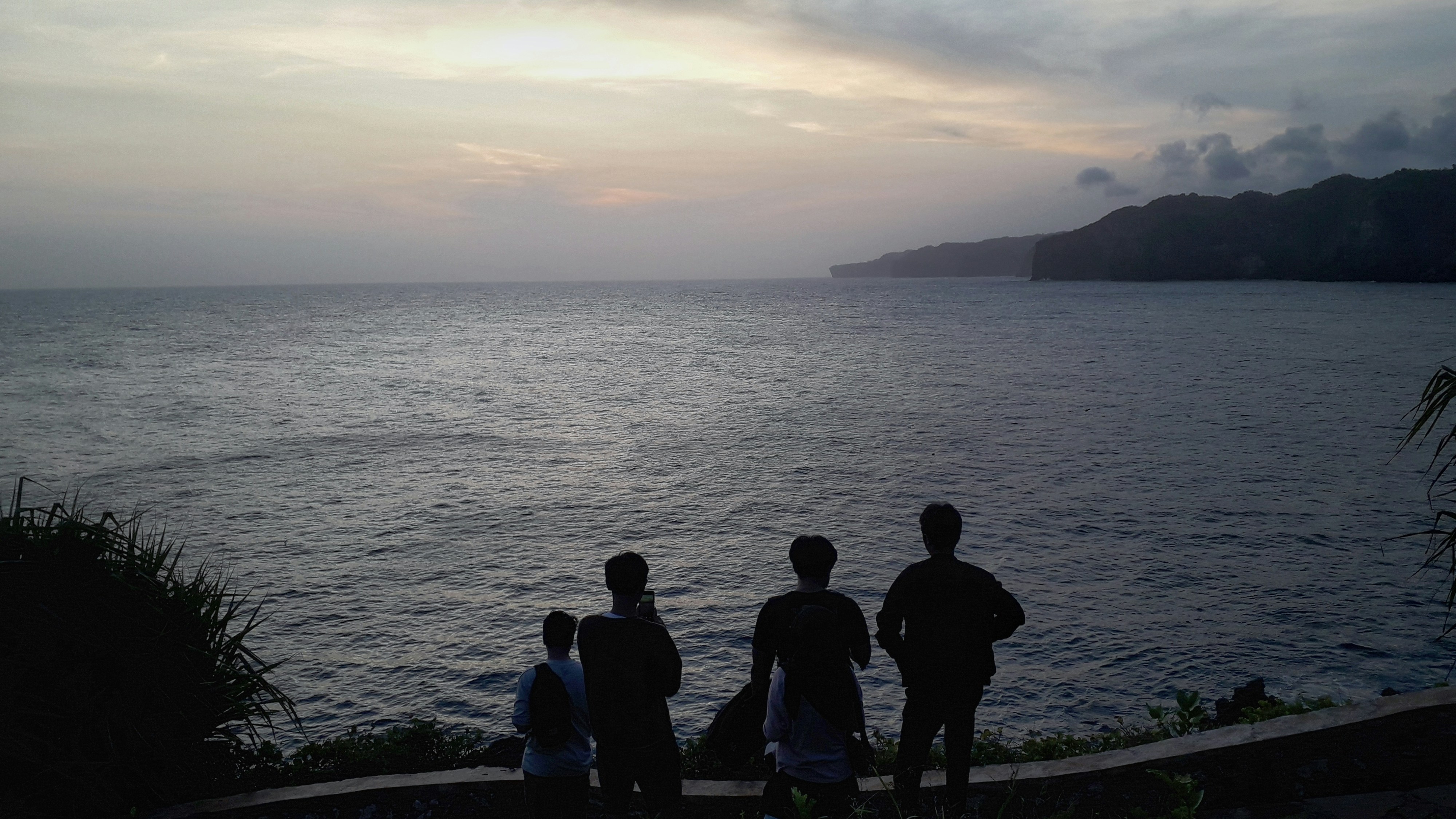 Four figures silhouetted against a tranquil sea at dusk, gazing at the horizon. The scene captures a serene moment of contemplation by the water's edge.
