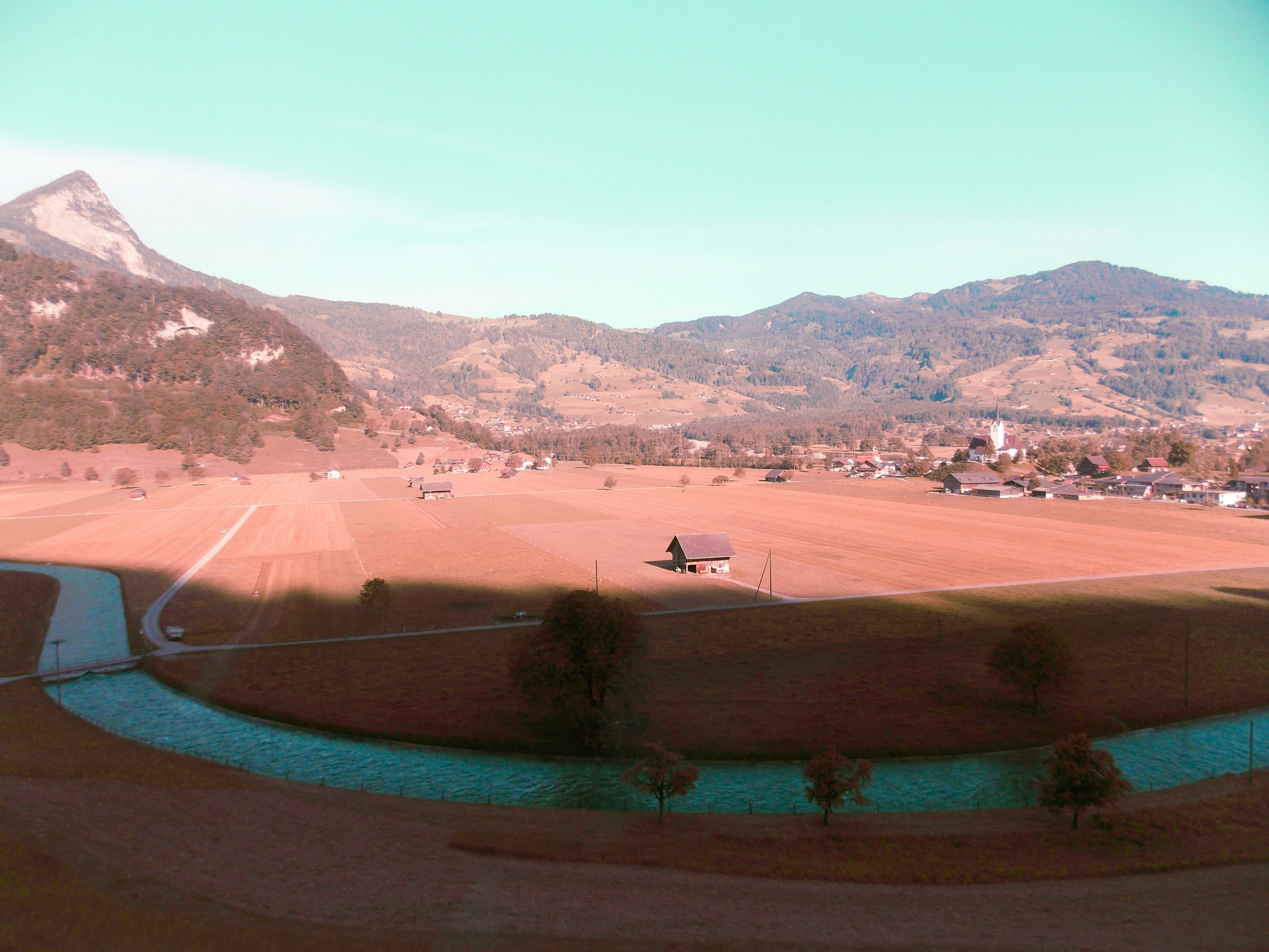 Expansive rural landscape featuring rolling fields, a winding river, and distant mountains under a pastel sky.