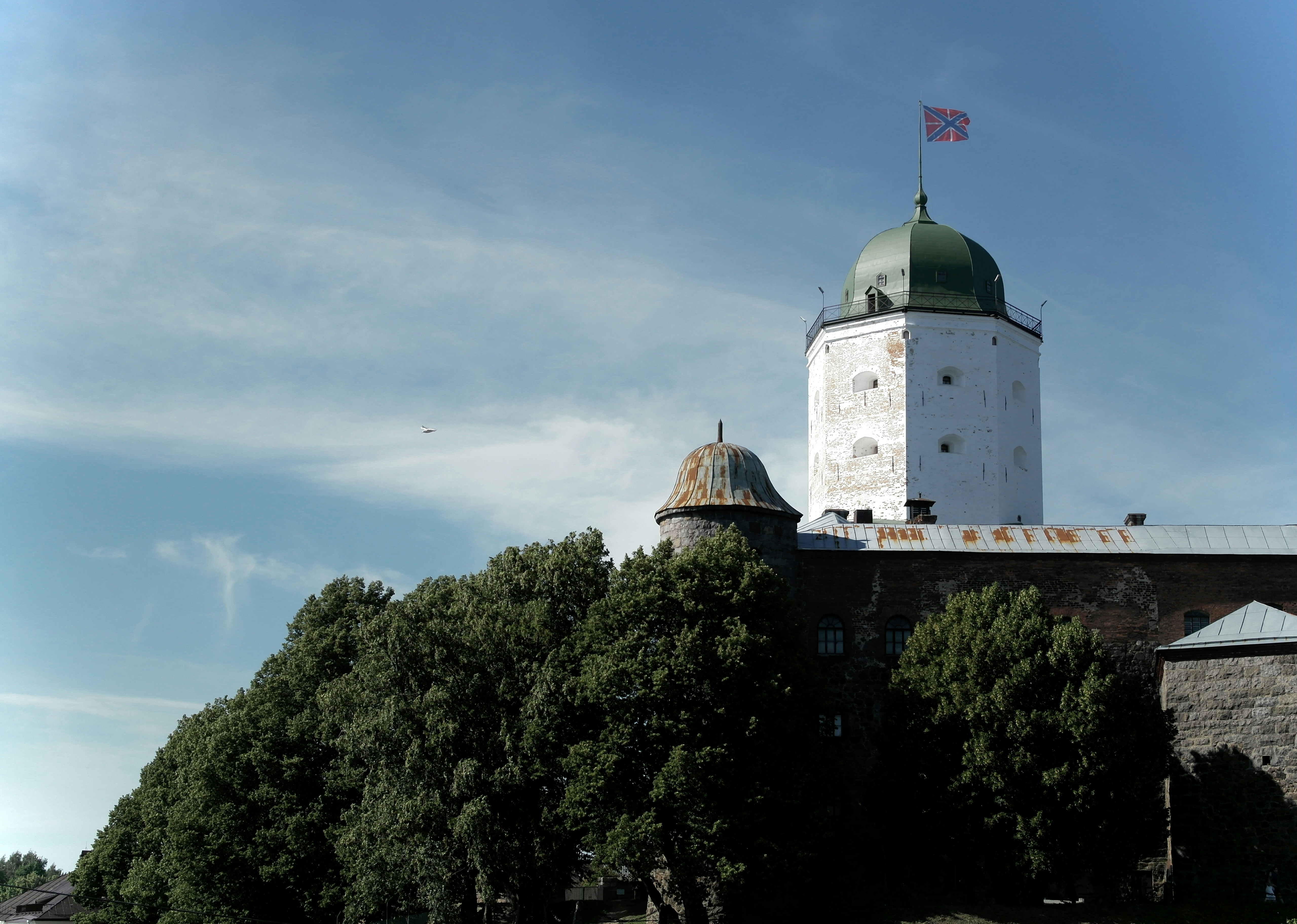 Historic fortress tower with a green dome and Union Jack flag, surrounded by lush trees under a bright sky.