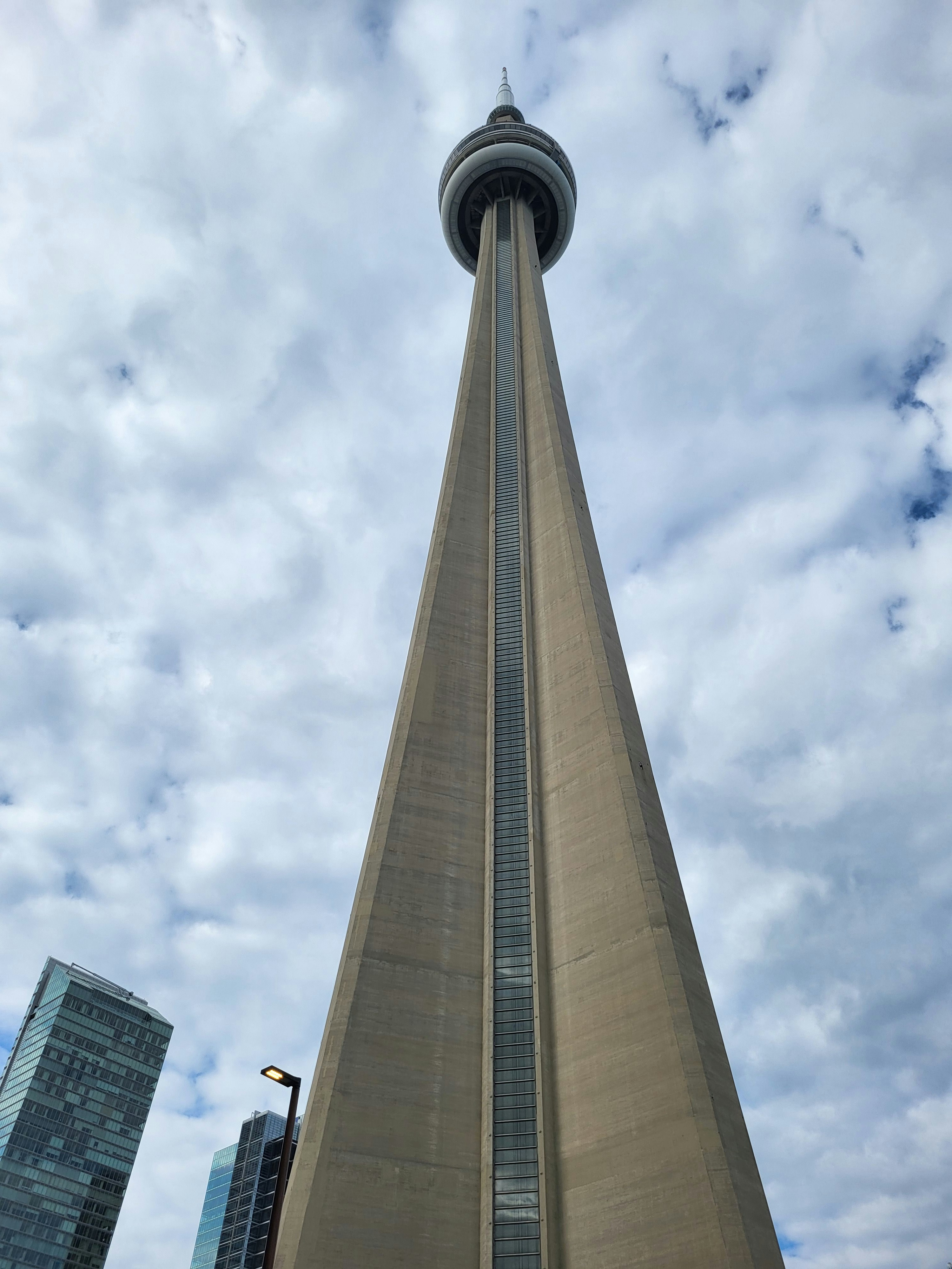 Upward view of the CN Tower against a backdrop of scattered clouds, flanked by modern skyscrapers.