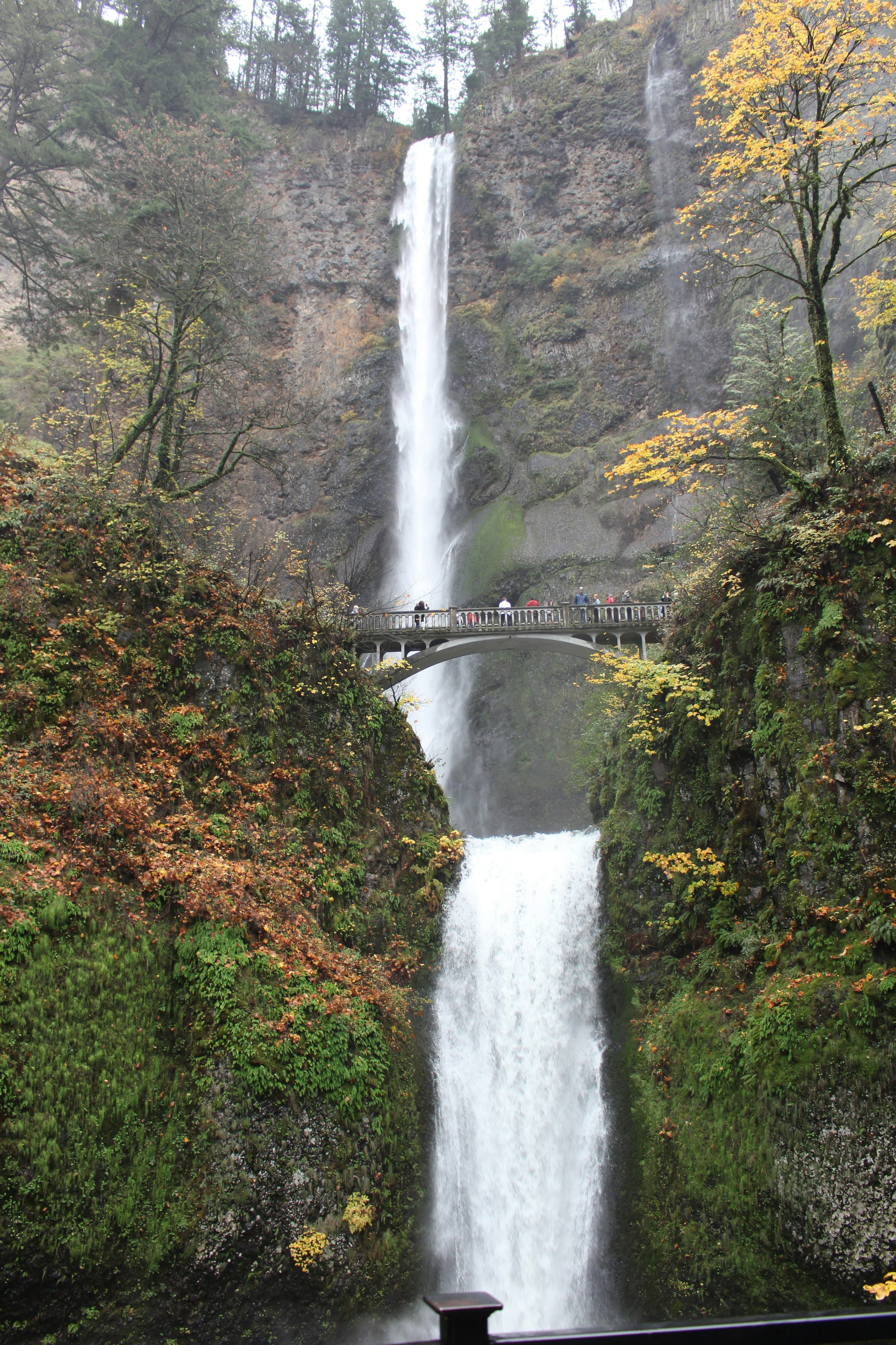 Majestic waterfall cascading down rocky cliffs, with a bridge connecting two sides amidst autumn foliage. Visitors can be seen enjoying the view.