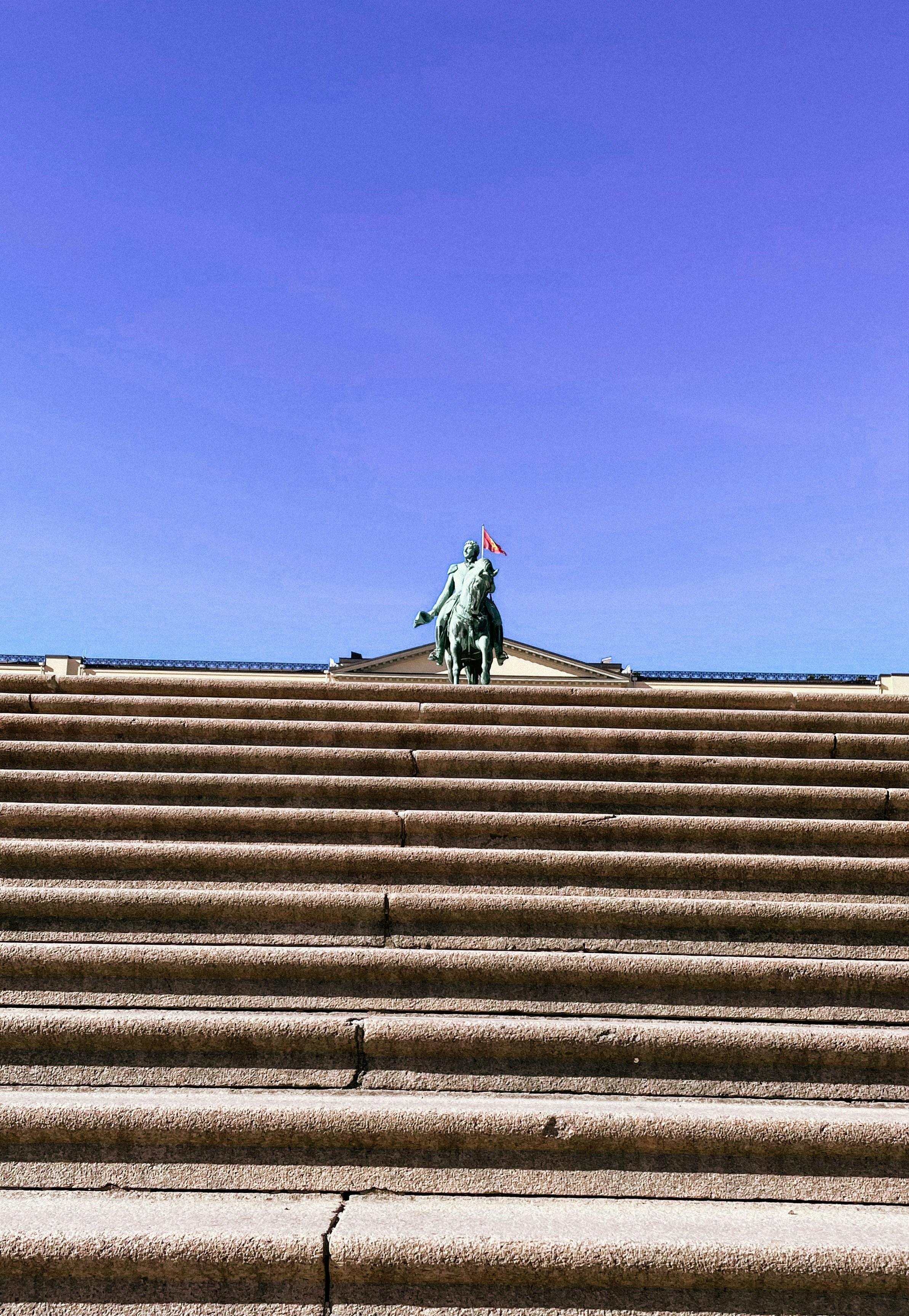 A grand staircase leading up to a historical monument, framed by a clear blue sky. The statue atop the stairs features a figure on horseback, adorned with a flag.