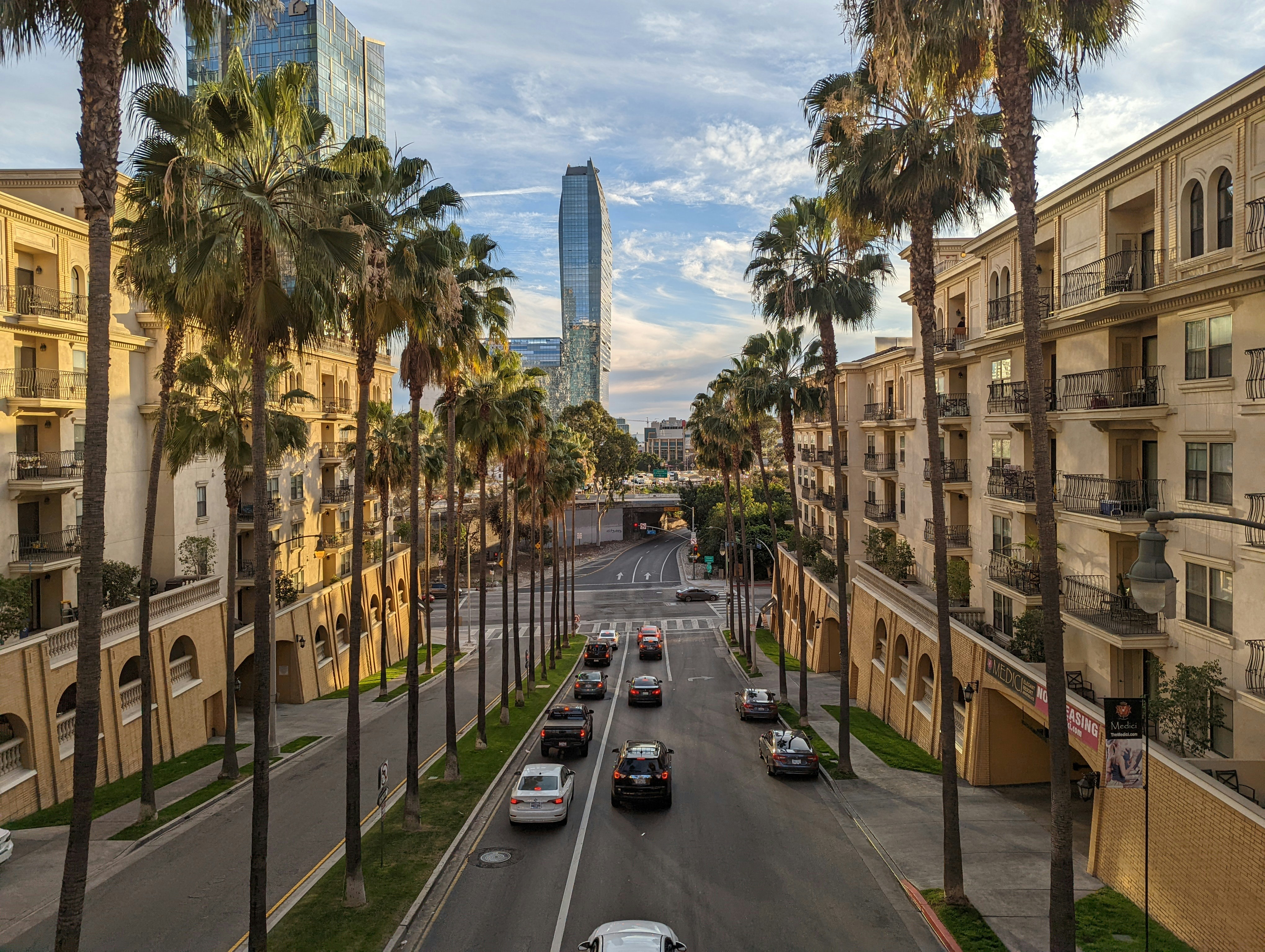 Palm trees line a bustling city street with a tall skyscraper in the distance under a partly cloudy sky.