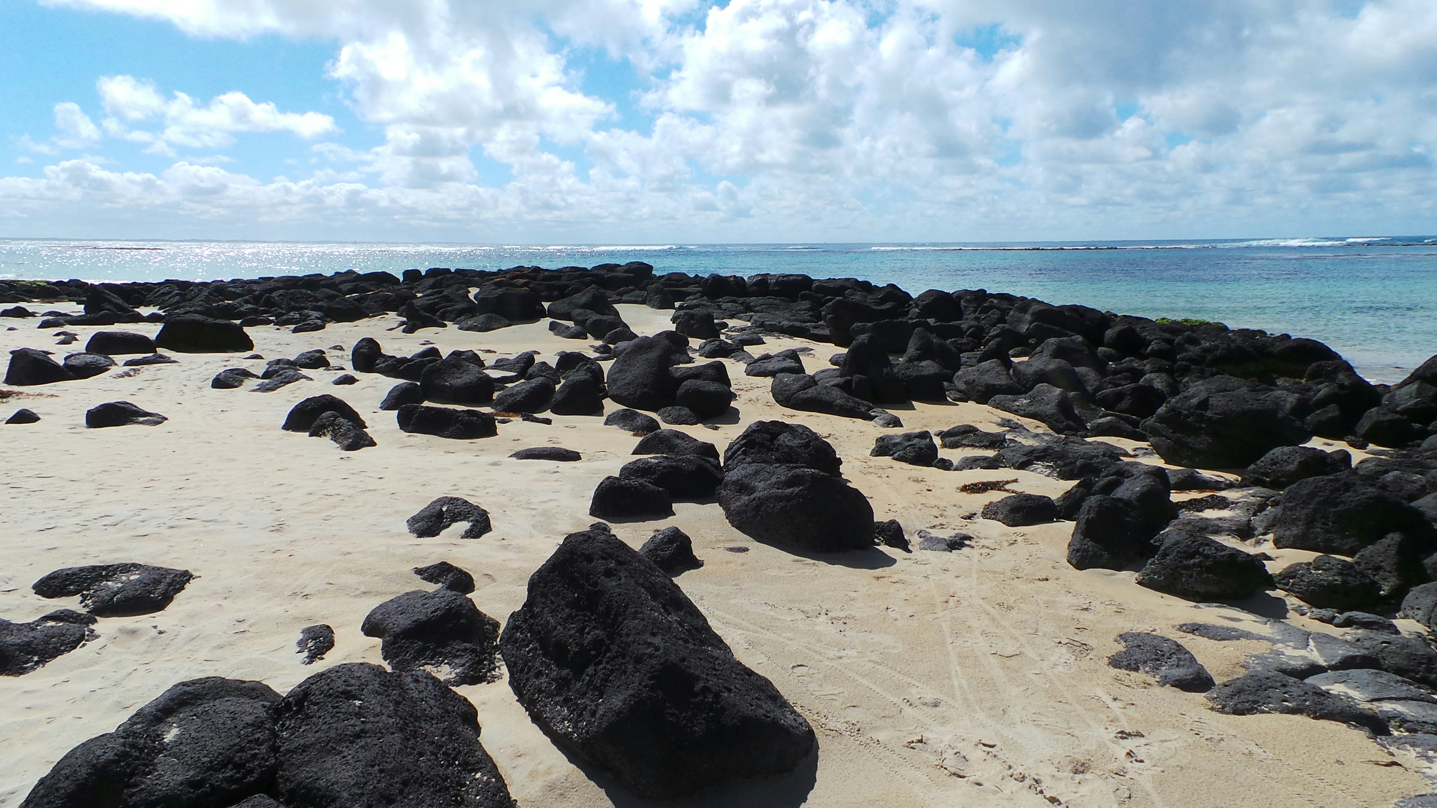 Wide seascape photograph featuring a sandy beach scattered with dark lava rocks, a calm turquoise sea, and a bright, cloud-filled sky.