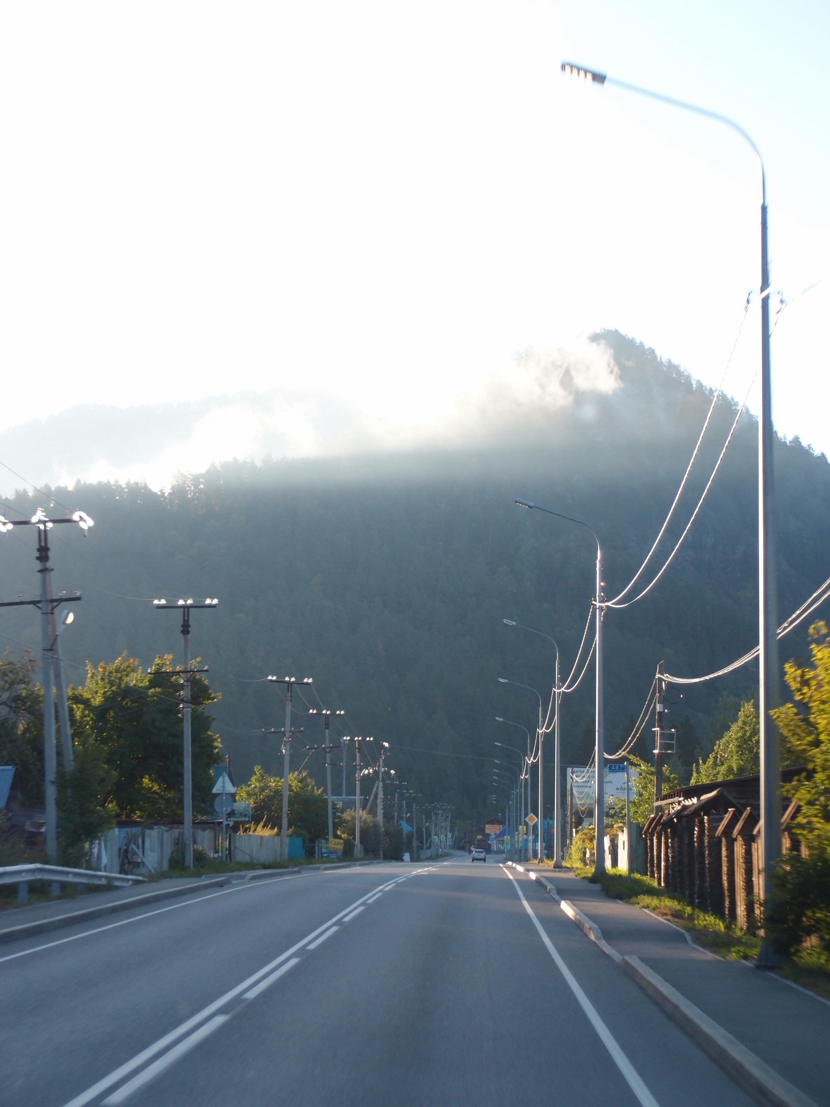 Quiet road stretches toward a forested hill, flanked by streetlights and utility poles as morning light floods the scene.