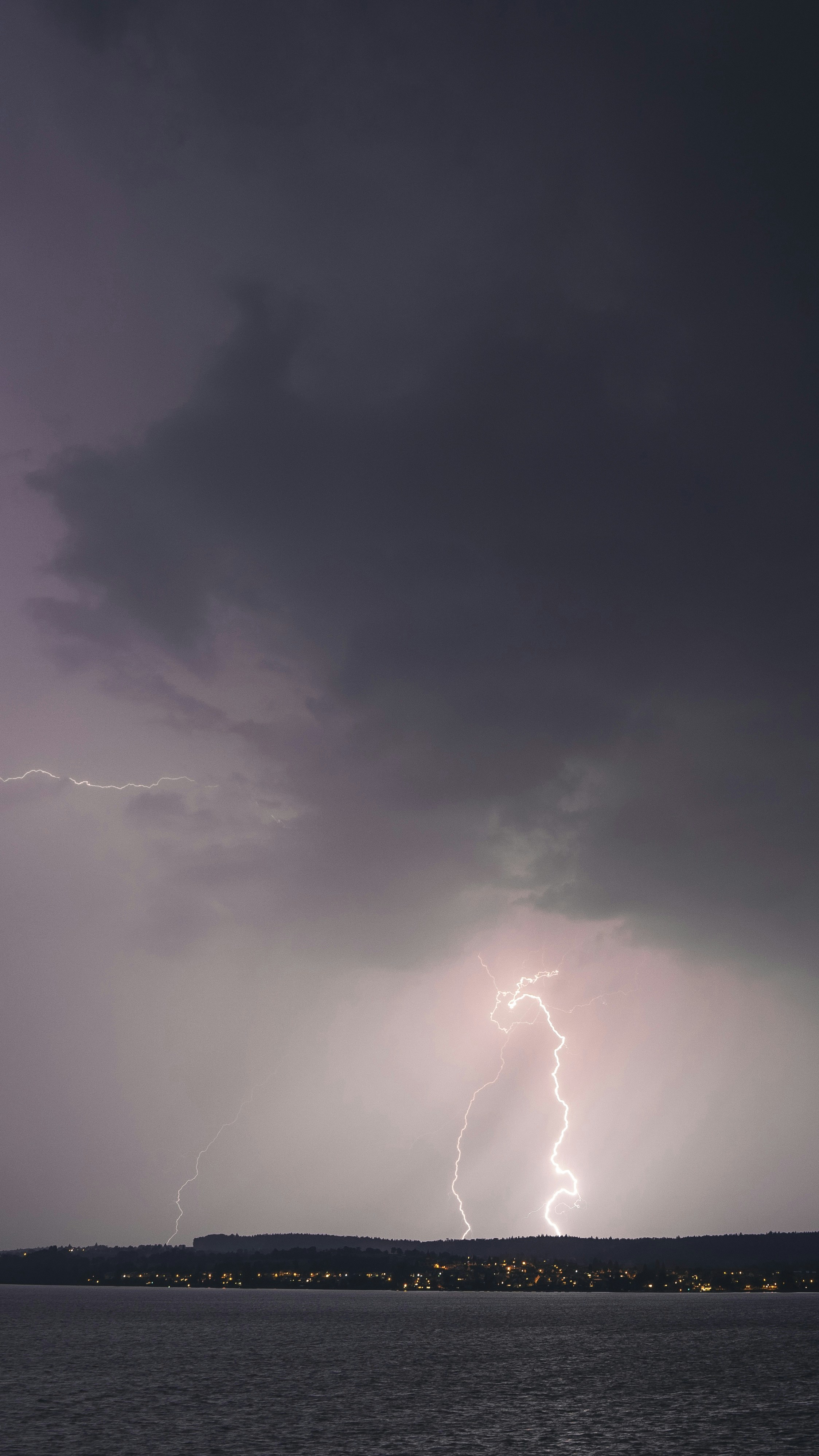 Lightning strikes dramatically illuminate the stormy sky above a tranquil body of water, with distant city lights twinkling below.