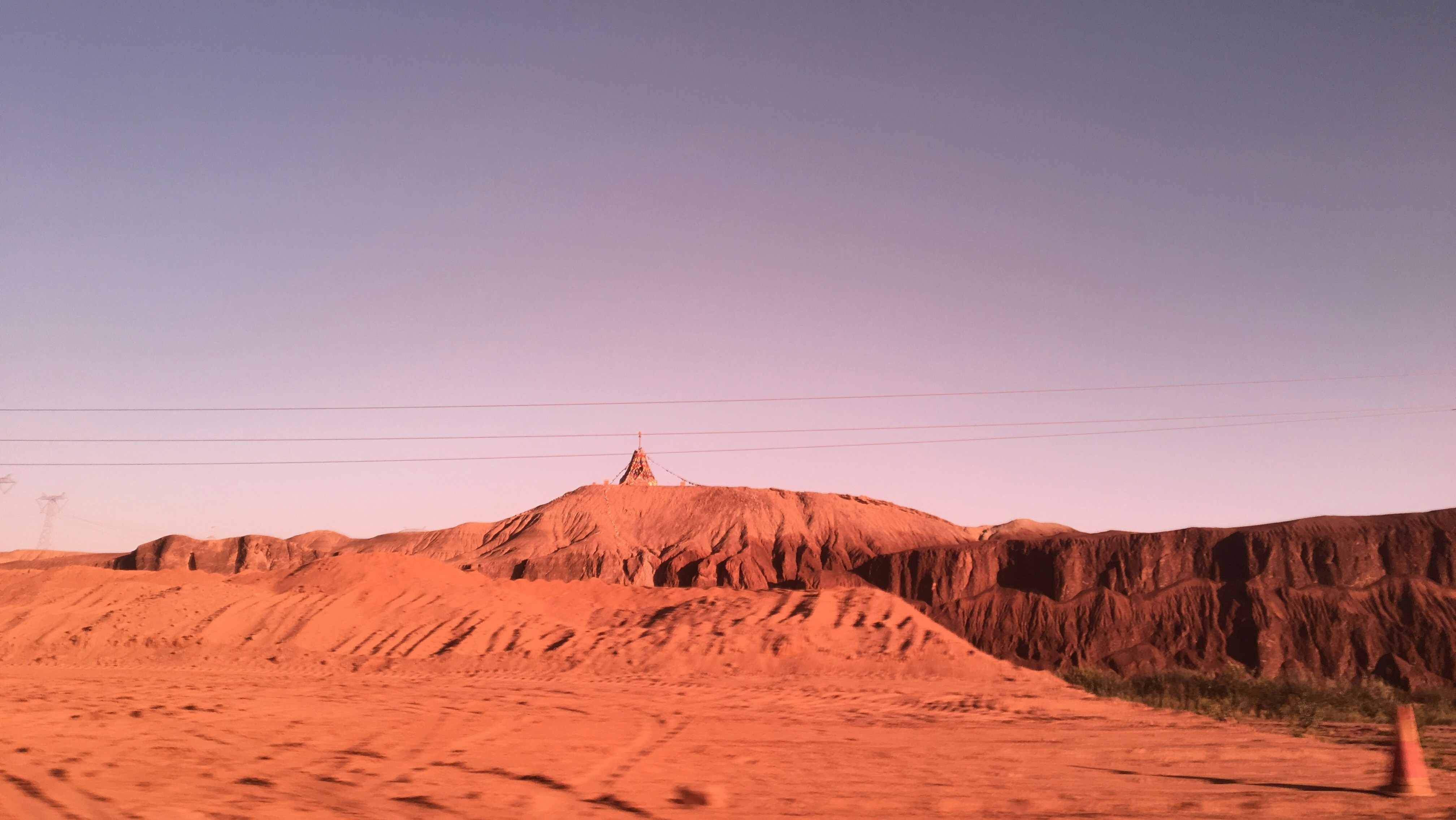 A vast expanse of reddish desert terrain with a prominent hill crowned by a structure, set against a gradient sky. Power lines stretch across the scene.