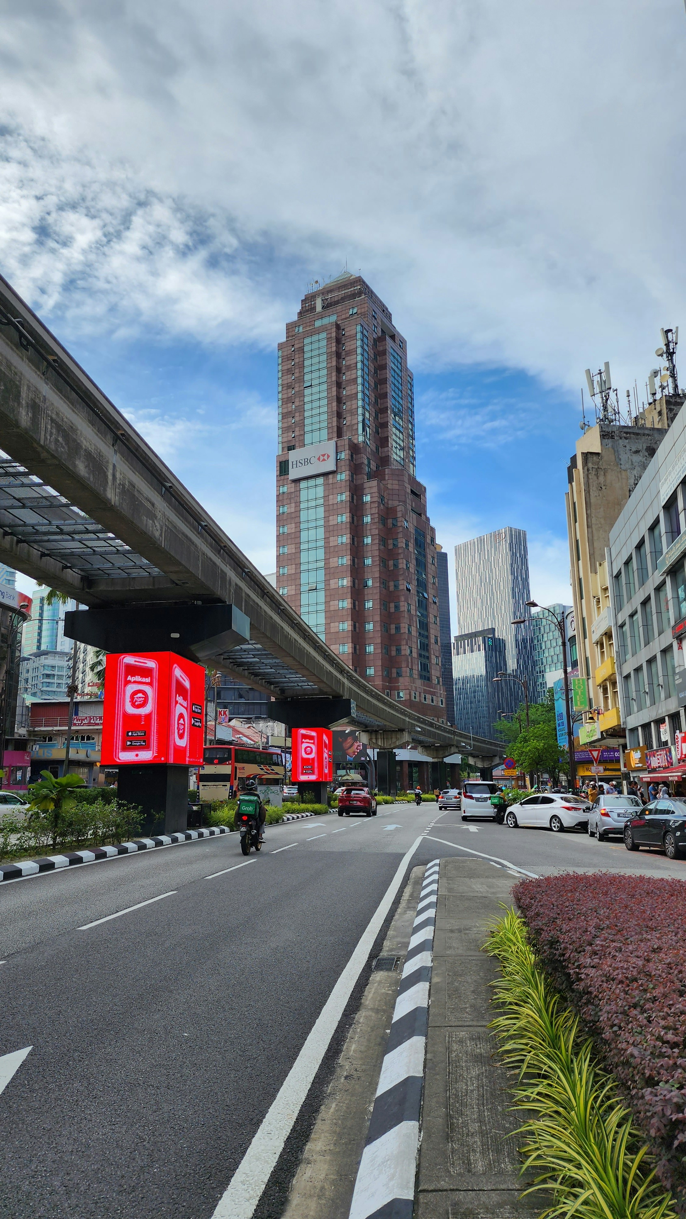 A busy urban street scene featuring a monorail above, flanked by modern skyscrapers and vibrant advertisements. The dynamic interplay of vehicles adds life to the cityscape.